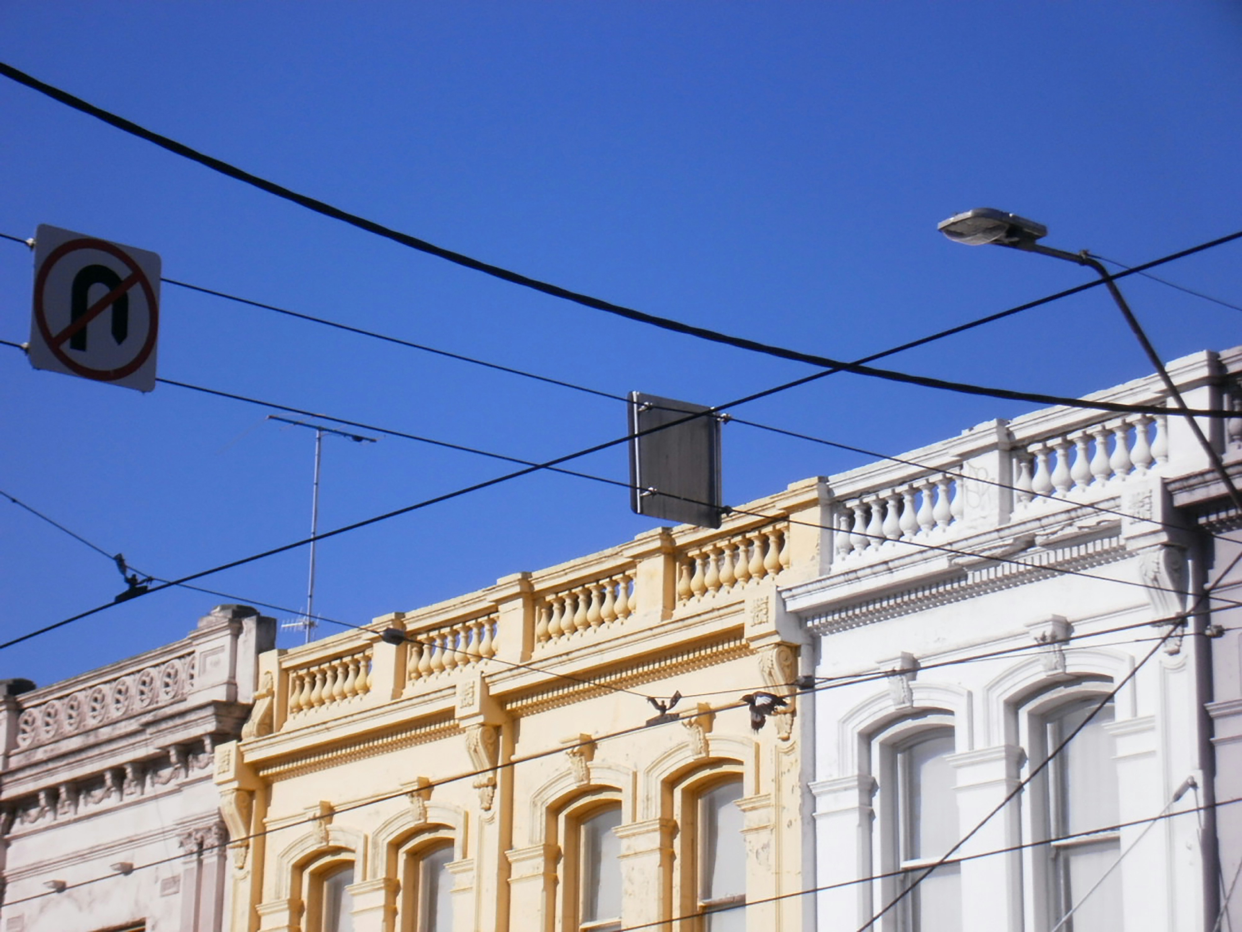 Historic building facades with ornate detailing beneath a network of overhead wires against a clear blue sky.