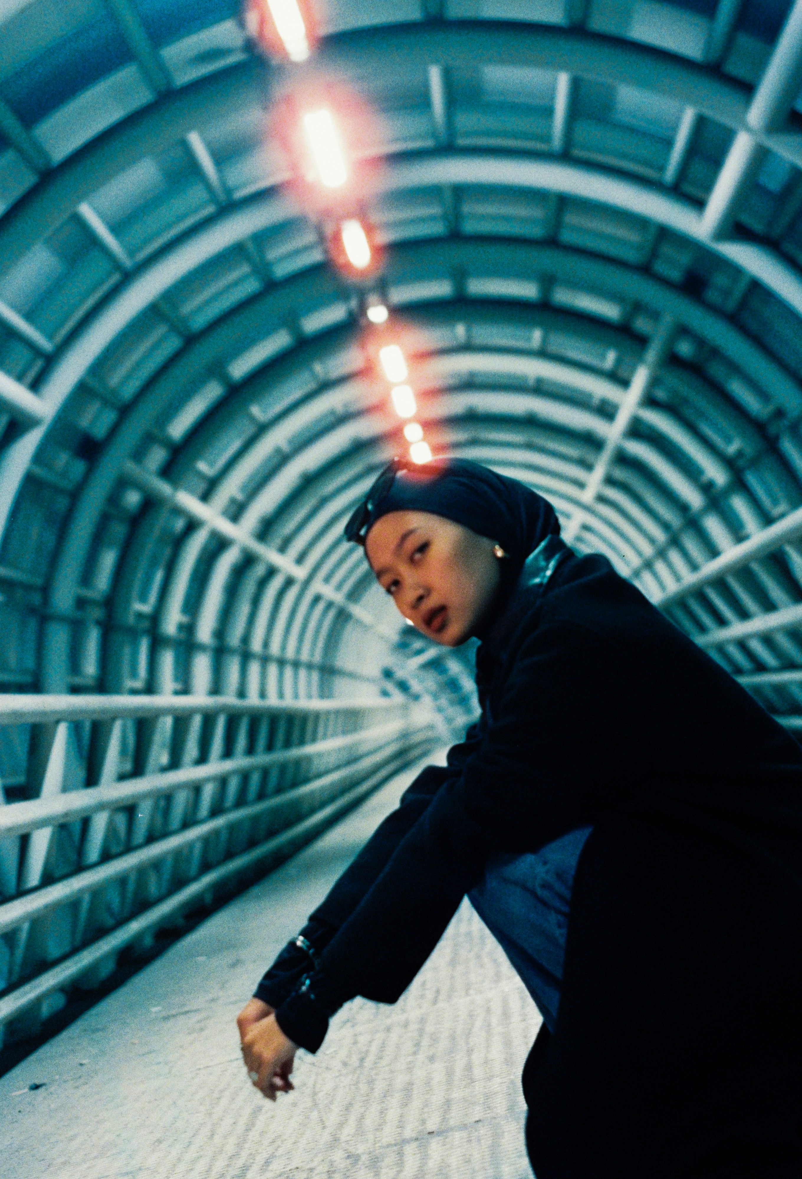 Woman crouching in a blue-hued, cylindrical tunnel with striking red light reflections along the ceiling.