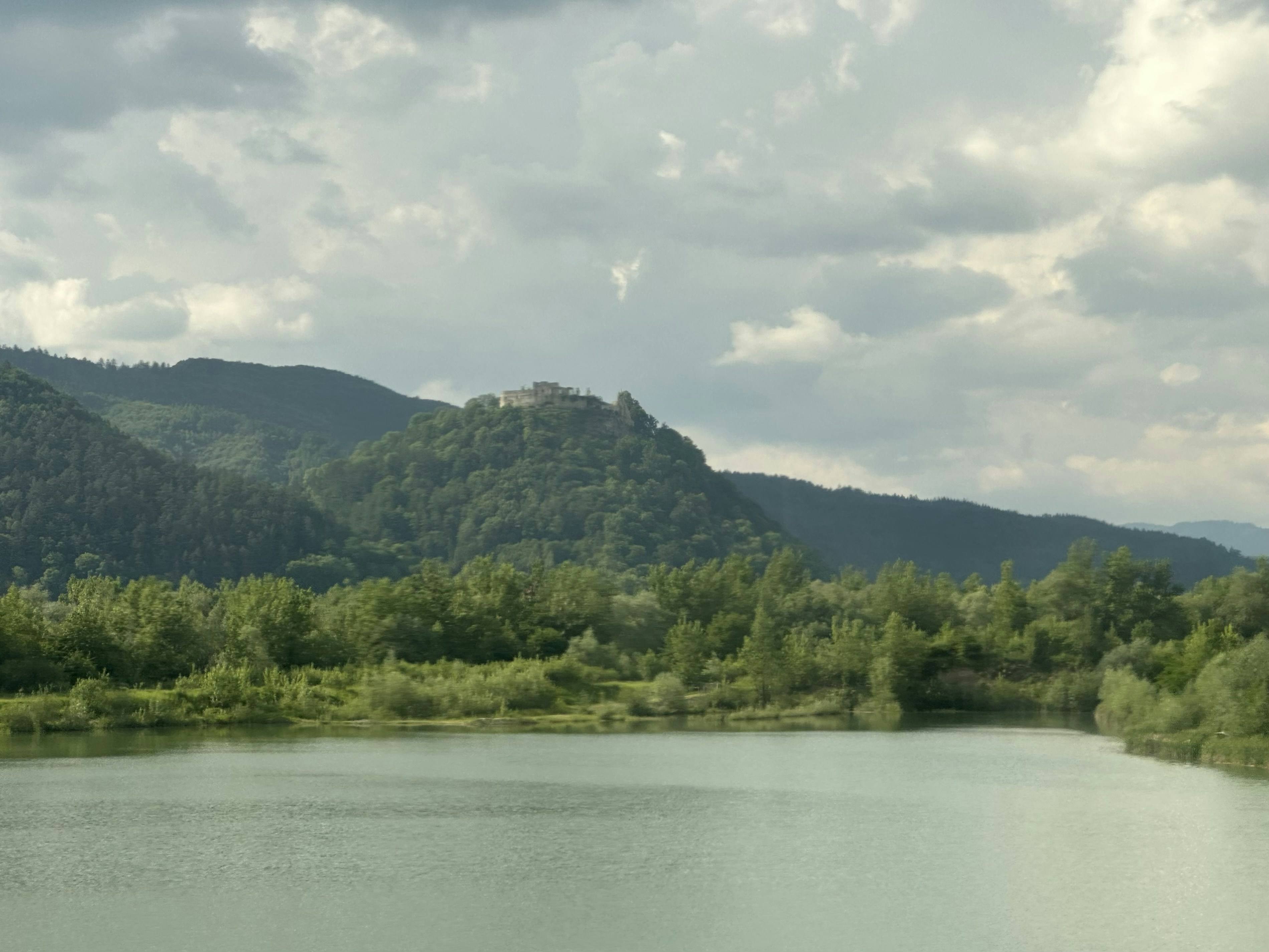 Calm river flowing through lush greenery with hills in the background under a cloudy sky.