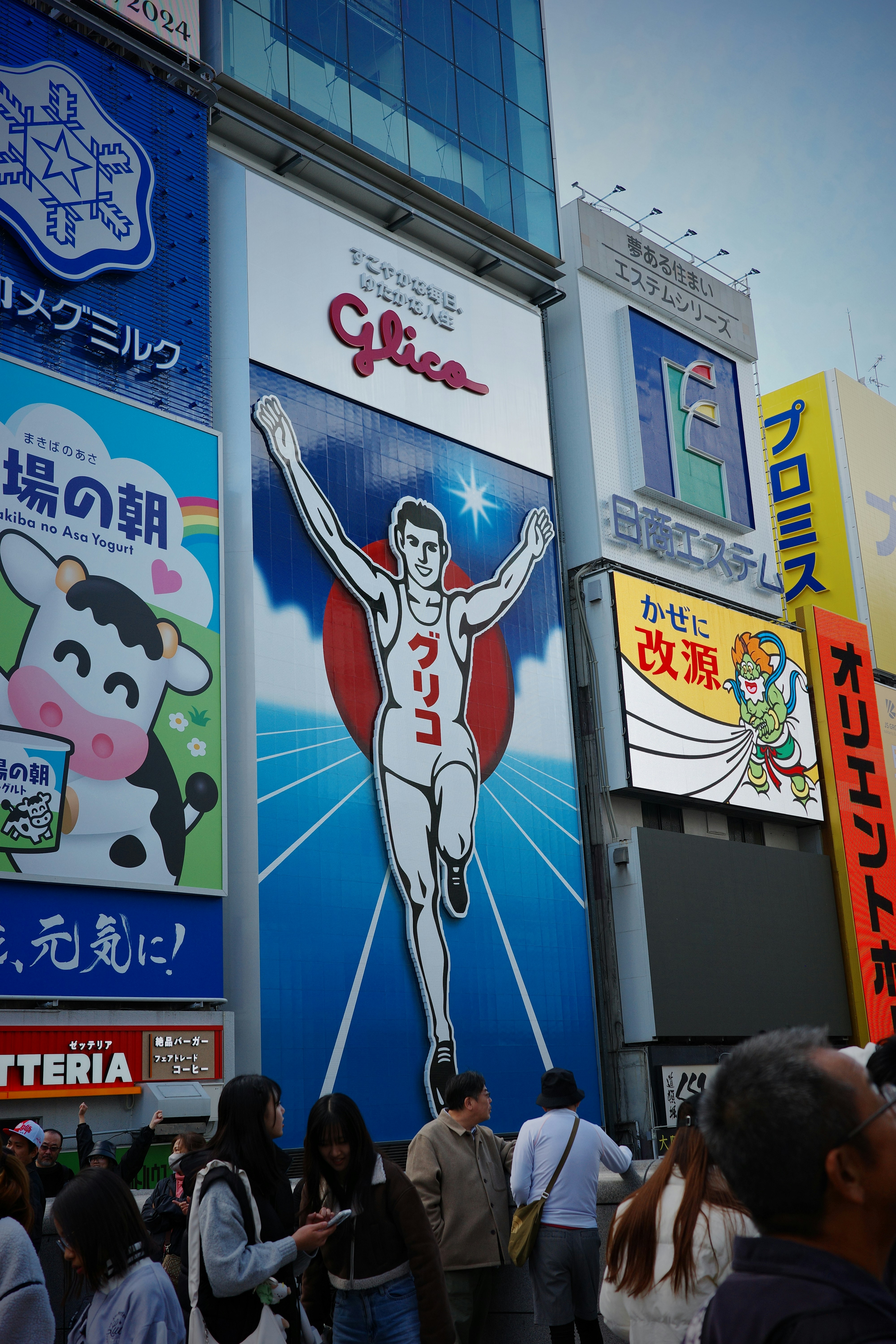 Vibrant advertisement featuring a runner with outstretched arms, set against a colorful backdrop of various billboards. The iconic Glico logo is prominently displayed.