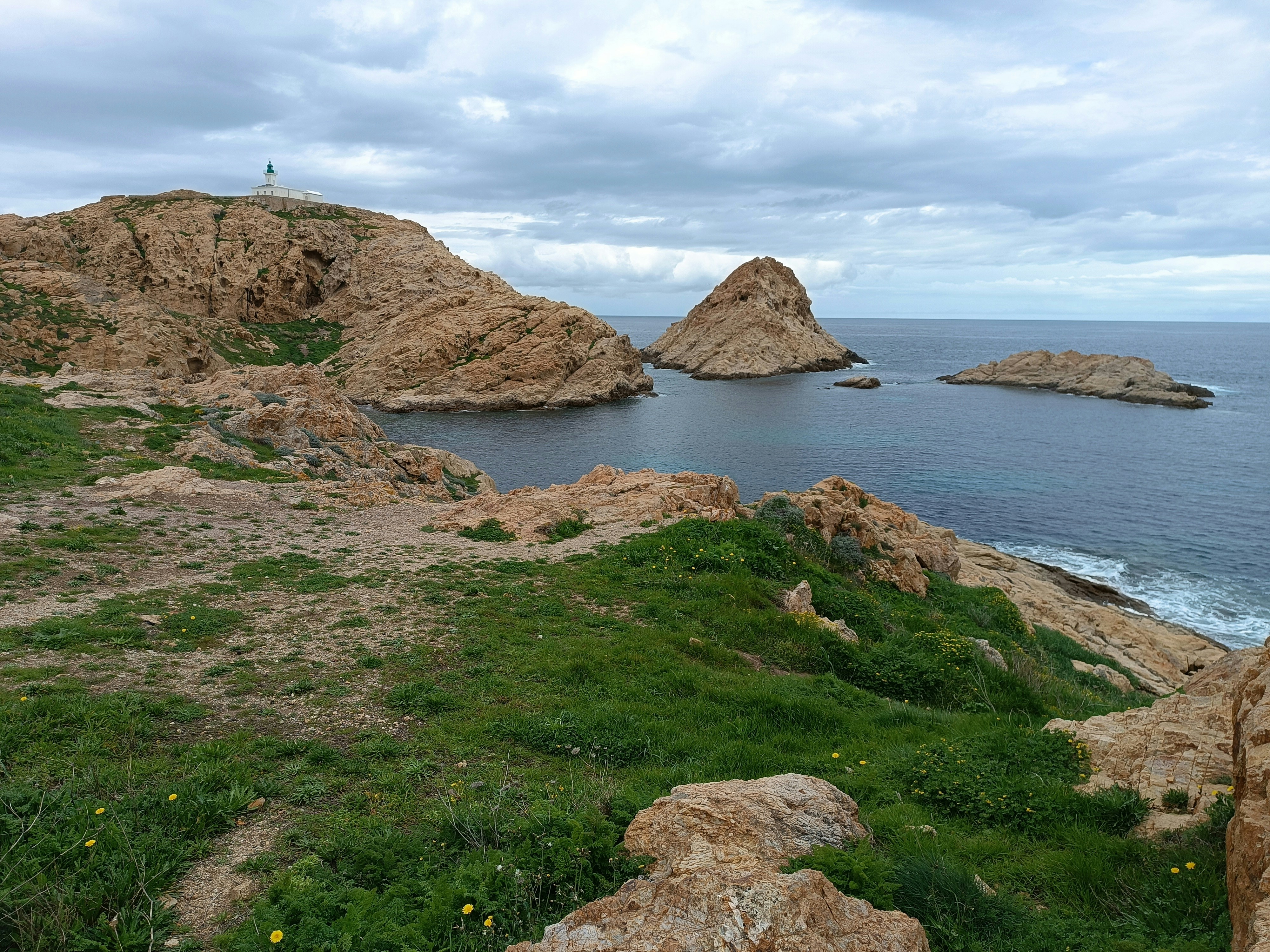 Coastal photograph showing rugged tan cliffs with a lighthouse on the left, green foreground, and a calm blue sea under a cloudy sky.