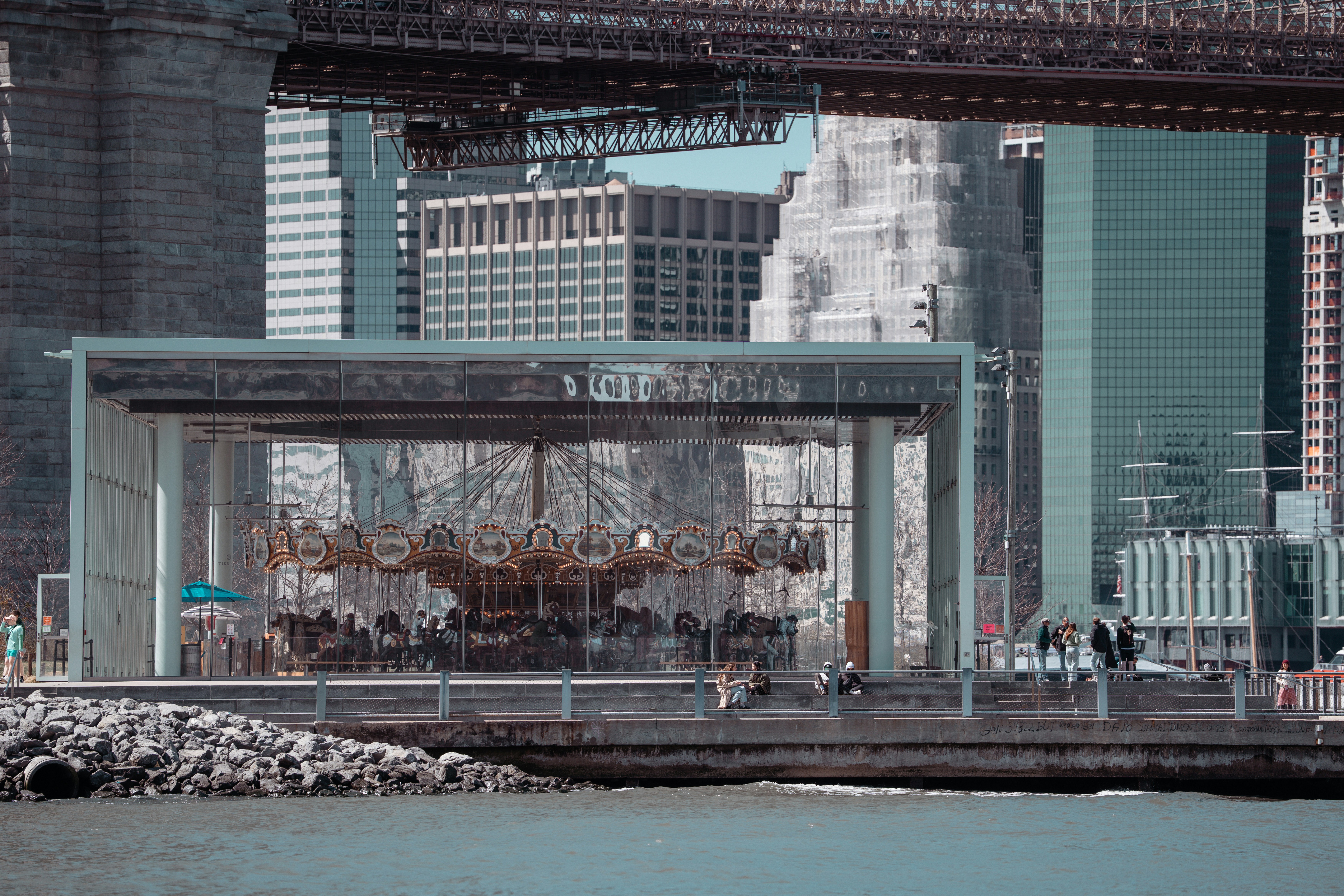 Glass-Pavilion Carousel Framed by a City Bridge and Skyline