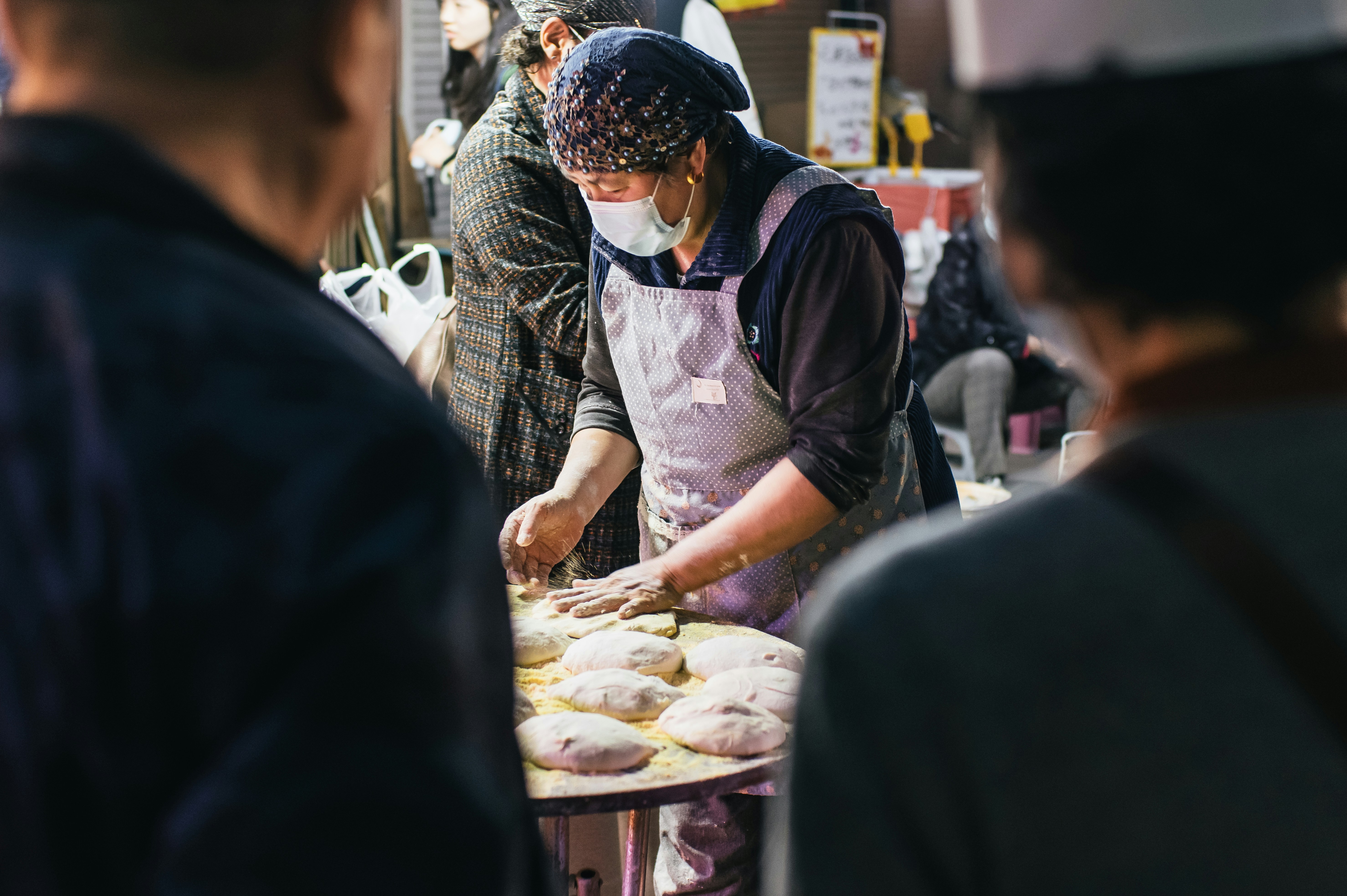 Masked baker in an apron shapes dough on a tray at a busy street market, with shoppers framing the scene.