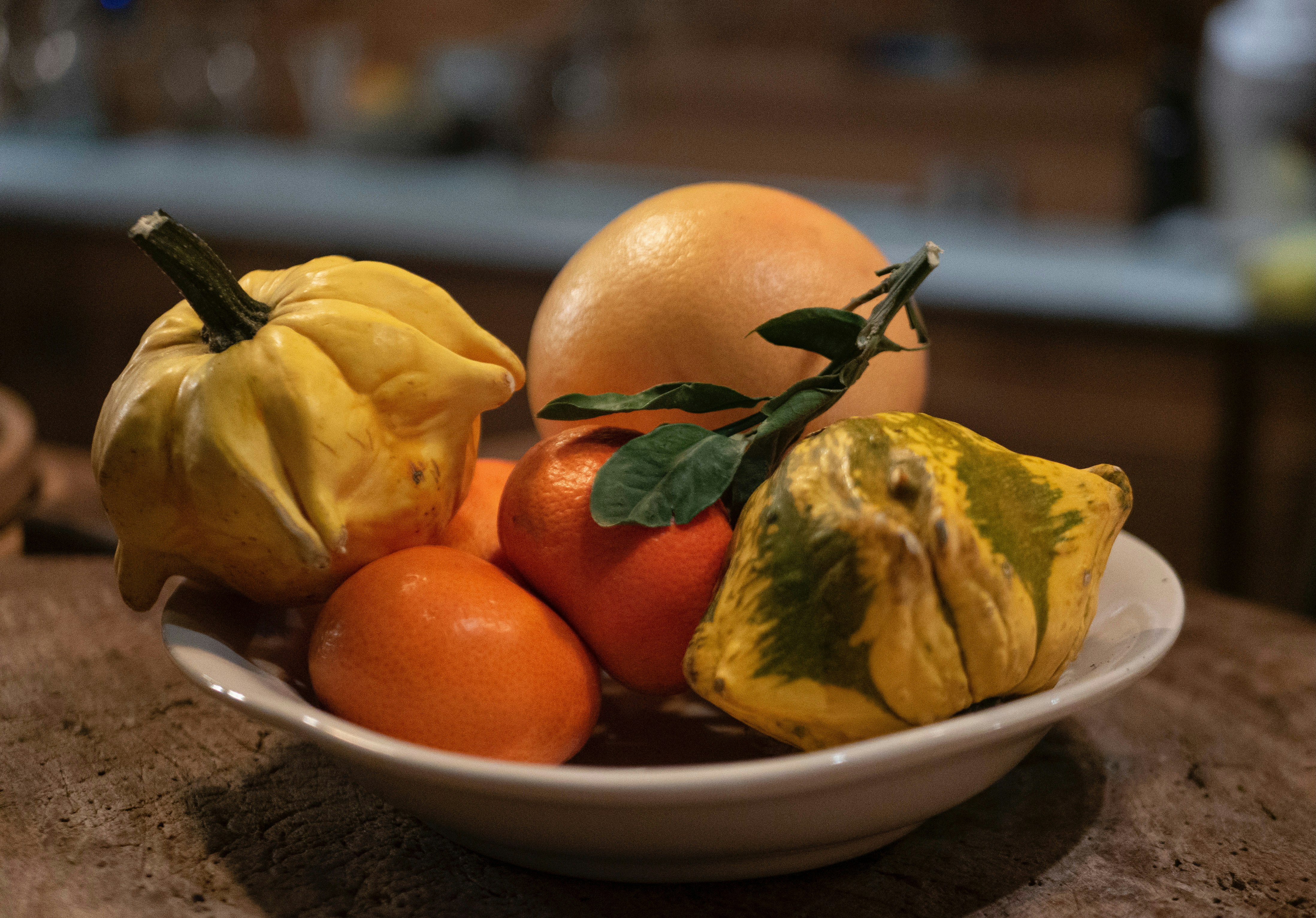 A plate of citrus fruits and gourds sits on a wooden surface, with warm ambient lighting highlighting the textures of the produce. The arrangement emphasizes natural colors and the rustic setting; note this is a still-life composition.