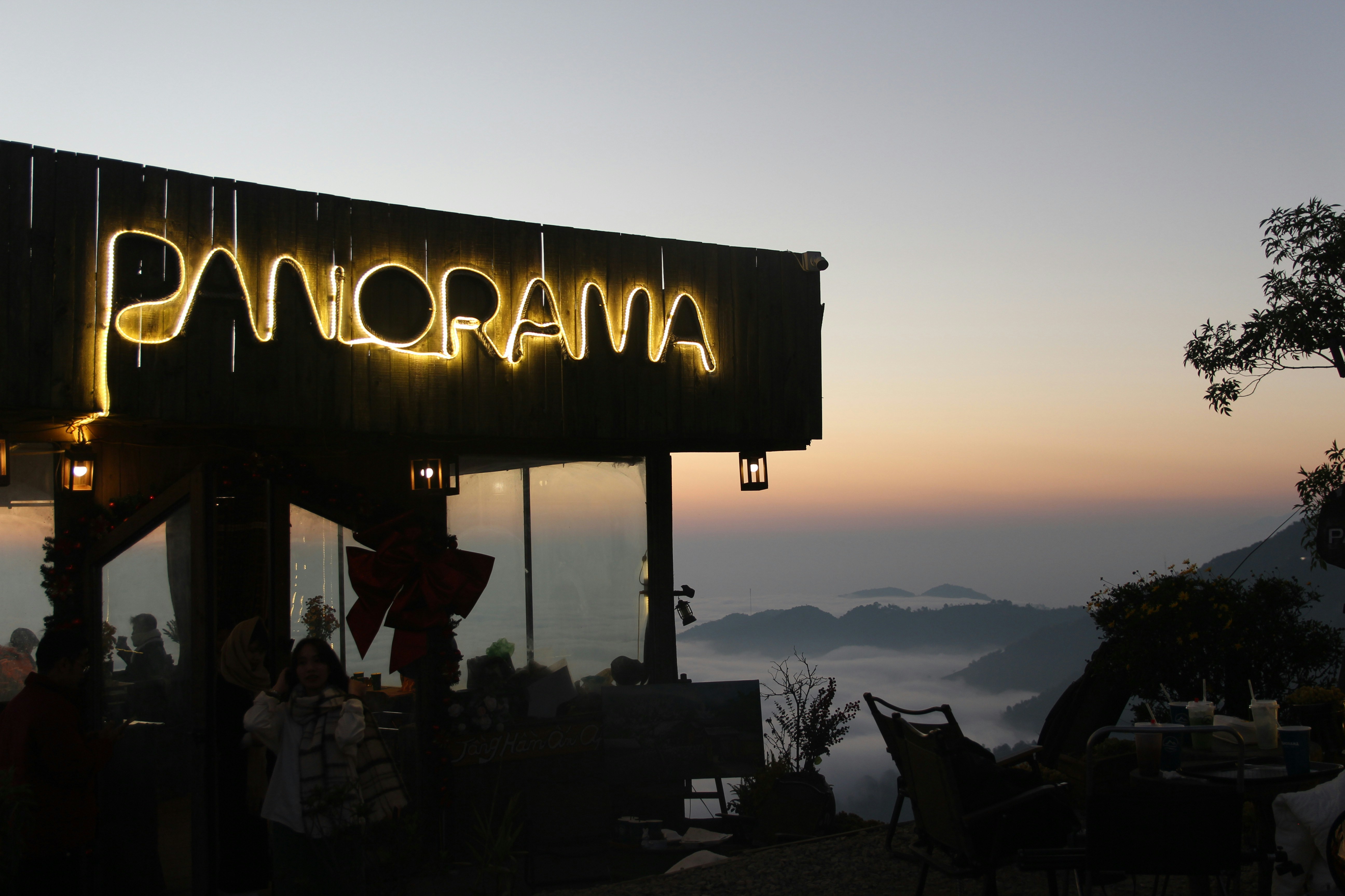 Silhouetted cafe with glowing 'Panorama' sign, overlooking misty mountains at sunset.