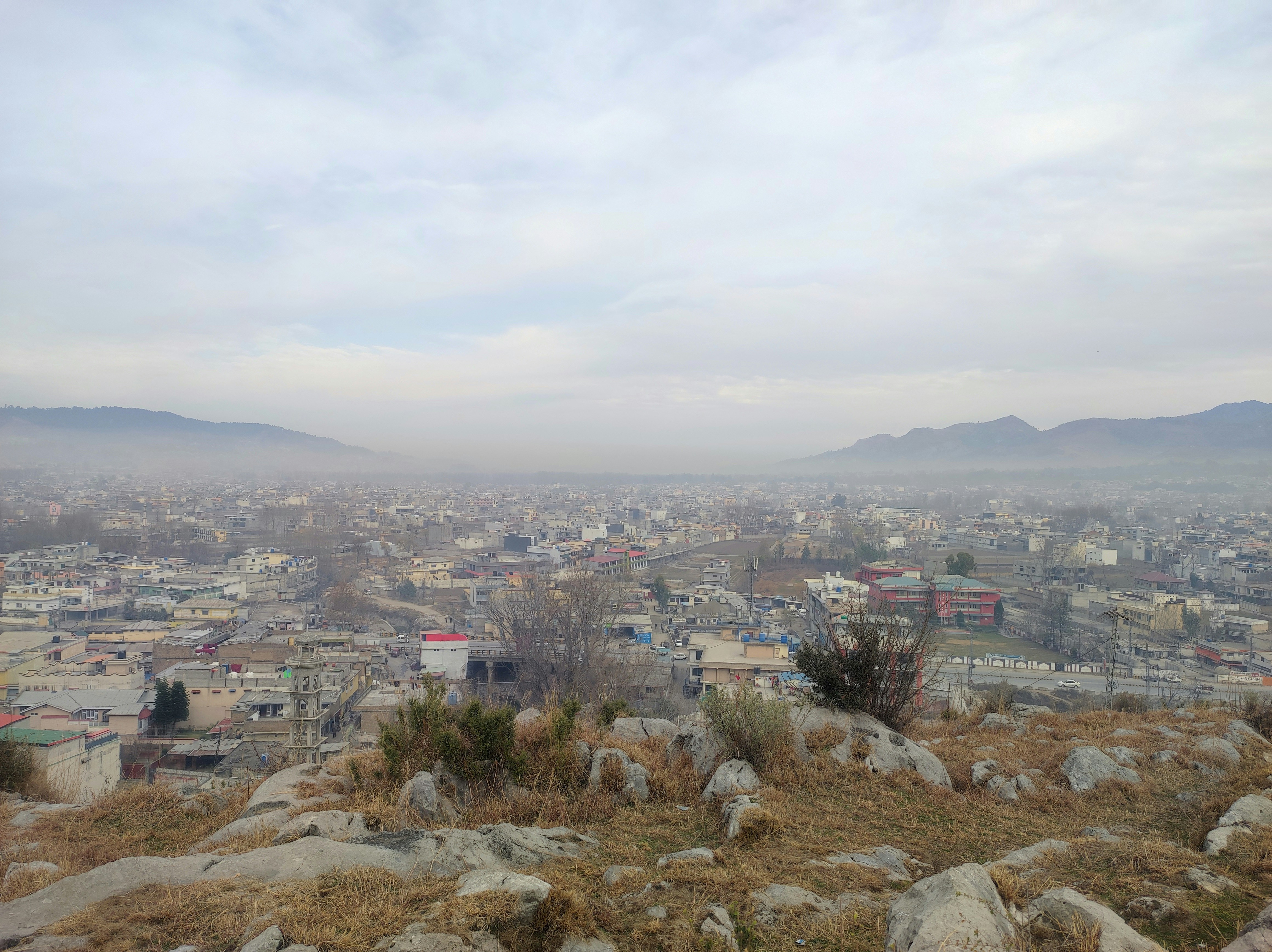 A panoramic view of a city shrouded in mist, with rocky terrain in the foreground and distant mountains fading into the haze.