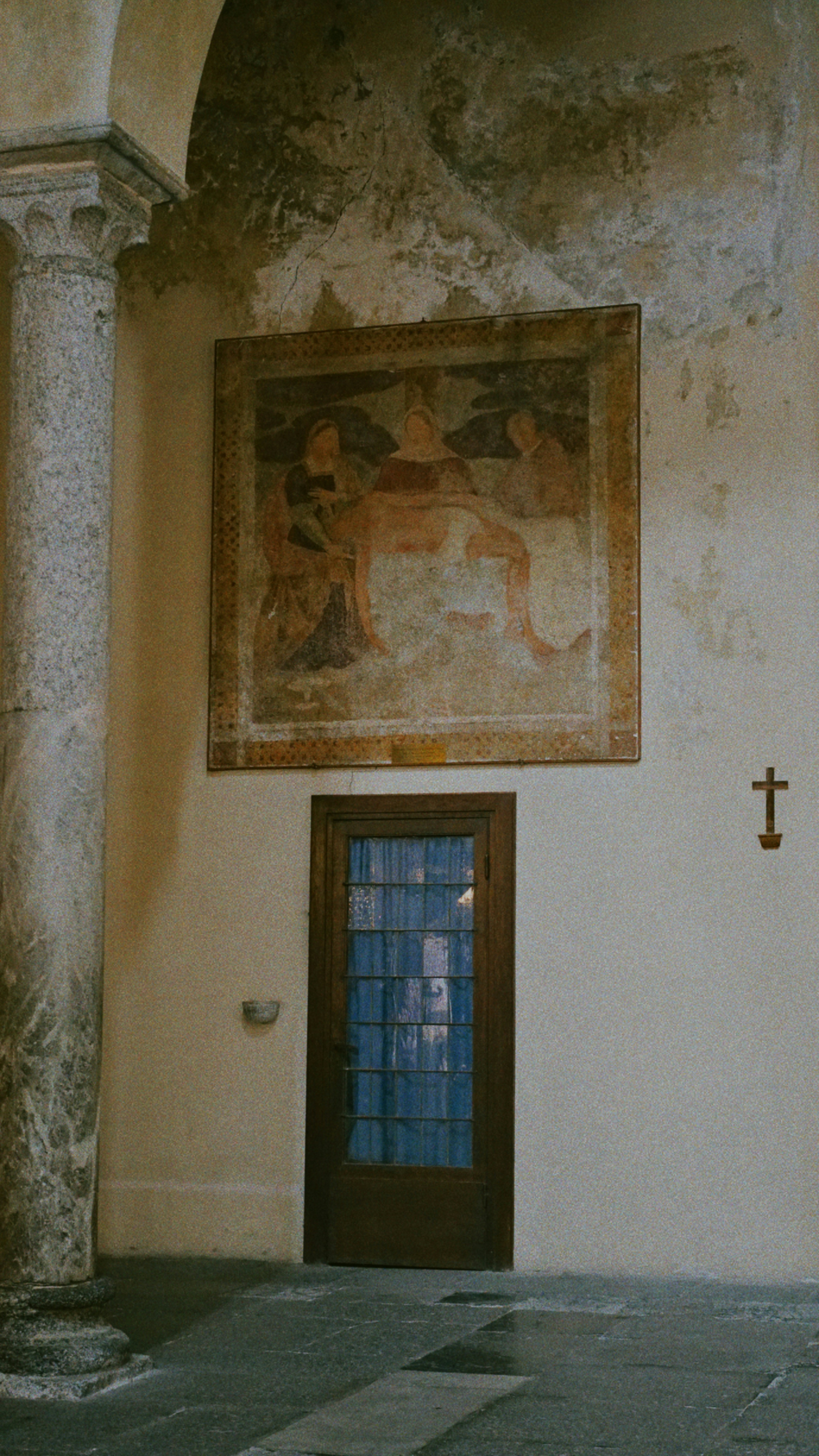 A rustic fresco adorns the wall above a wooden door in a dimly lit stone alcove.