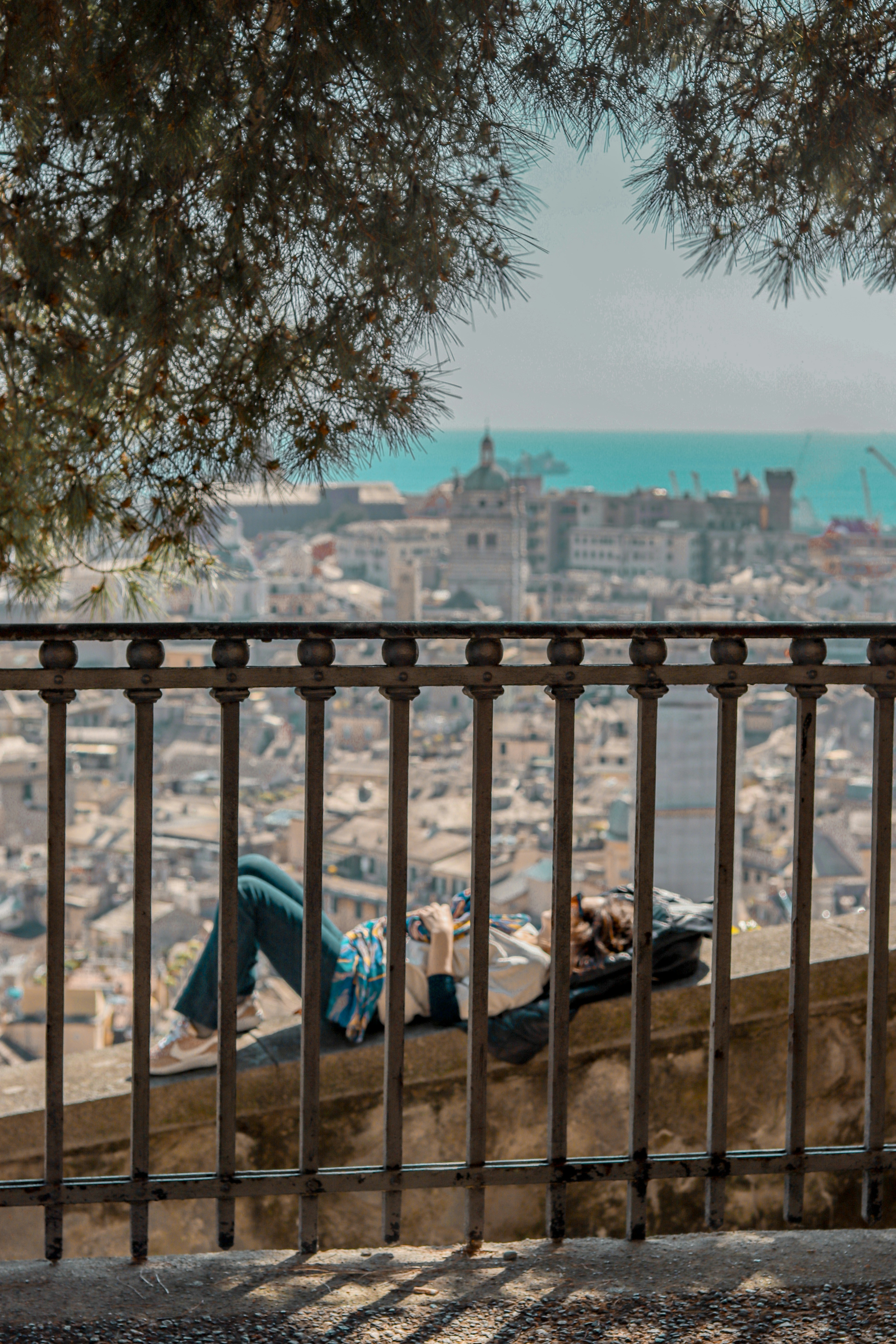 Person relaxing on a stone ledge overlooking a cityscape with a distant view of the sea.