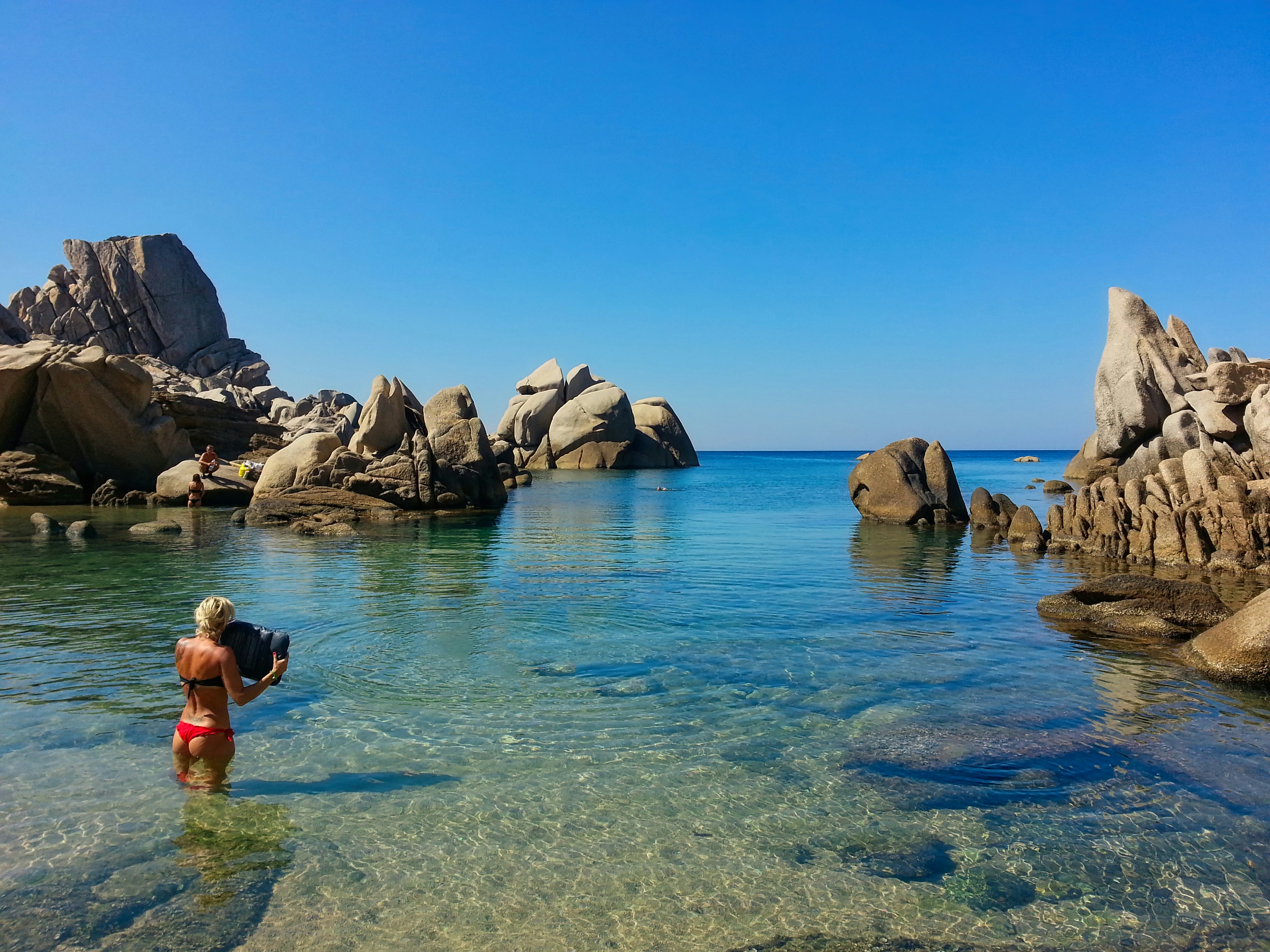 Person standing in clear, shallow water surrounded by jagged rock formations under a bright blue sky.