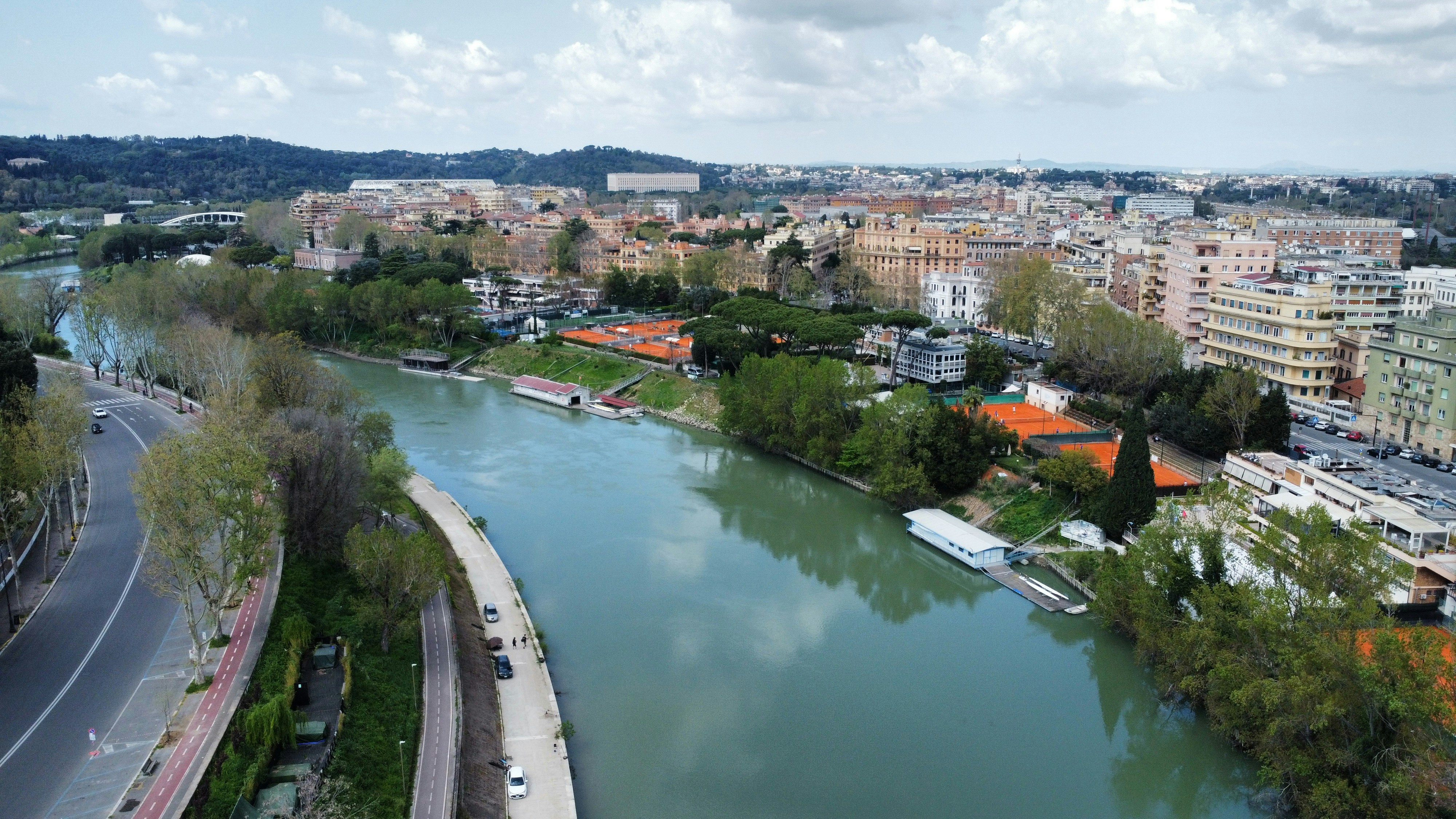 Aerial view of a river winding through a cityscape with surrounding greenery and buildings under a cloudy sky.