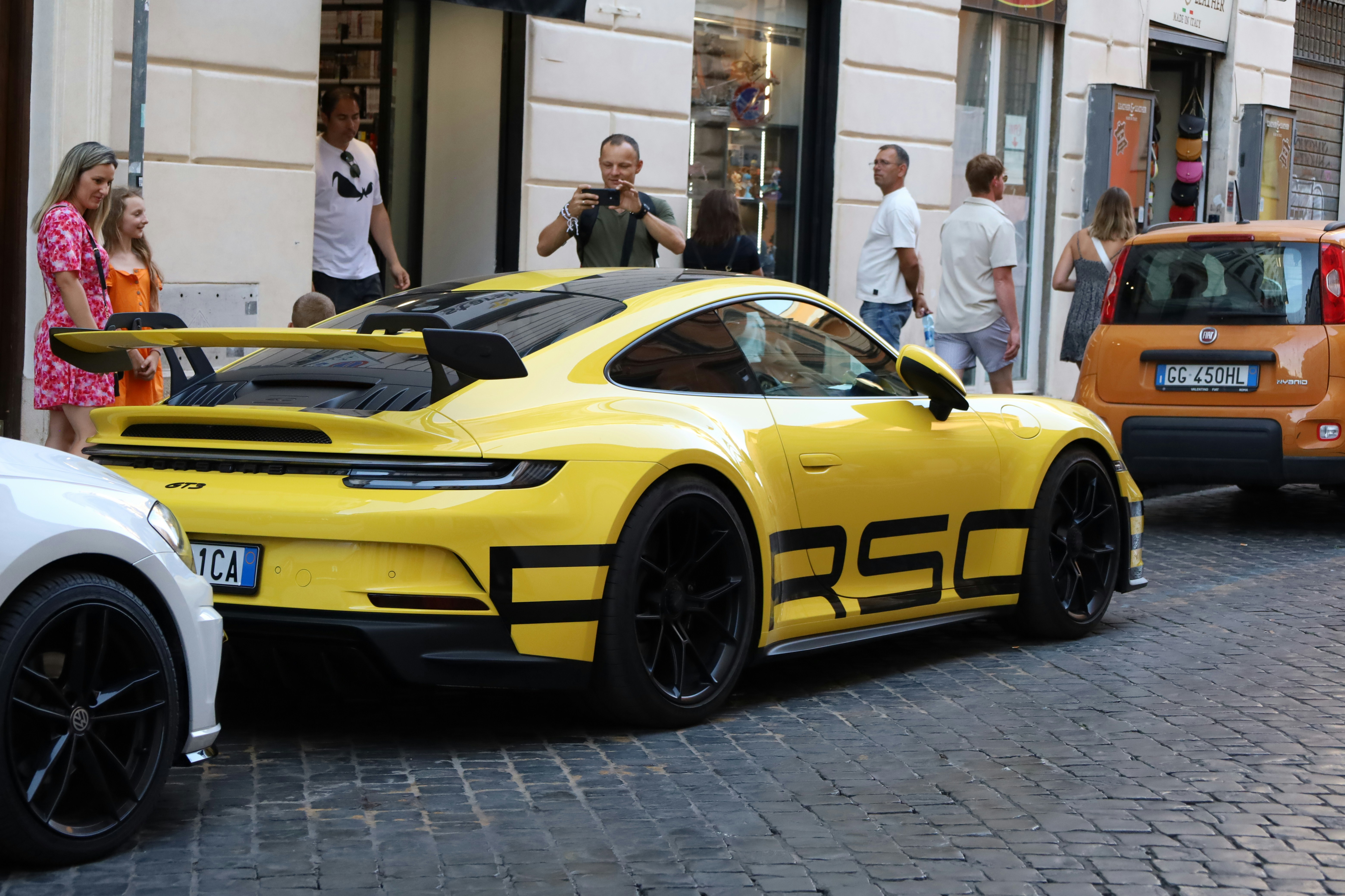 Yellow sports car with bold black detailing parked on a cobblestone street, surrounded by pedestrians and buildings.