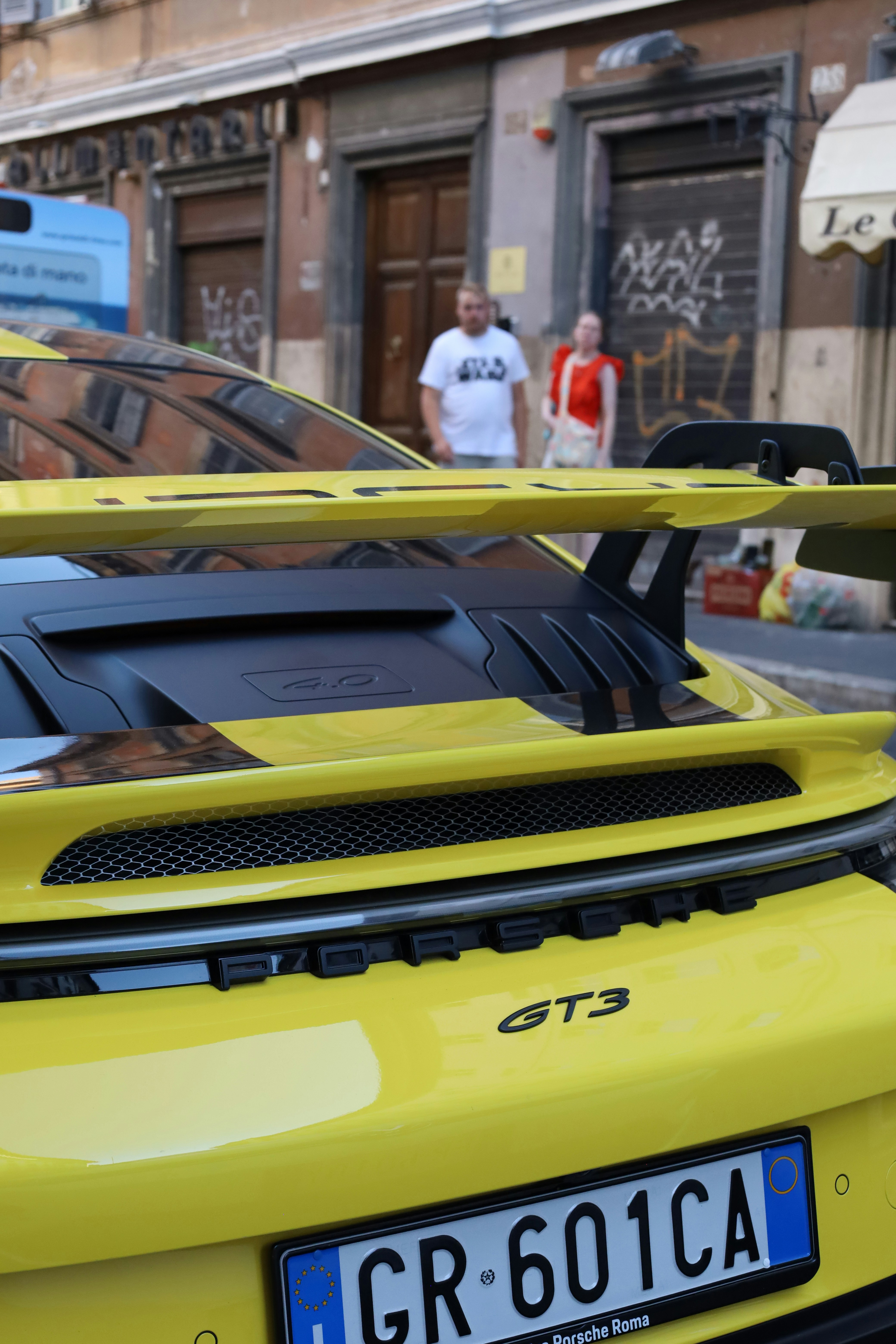 Yellow sports car with GT3 logo parked on a city street, with graffiti-covered walls and pedestrians in the background.