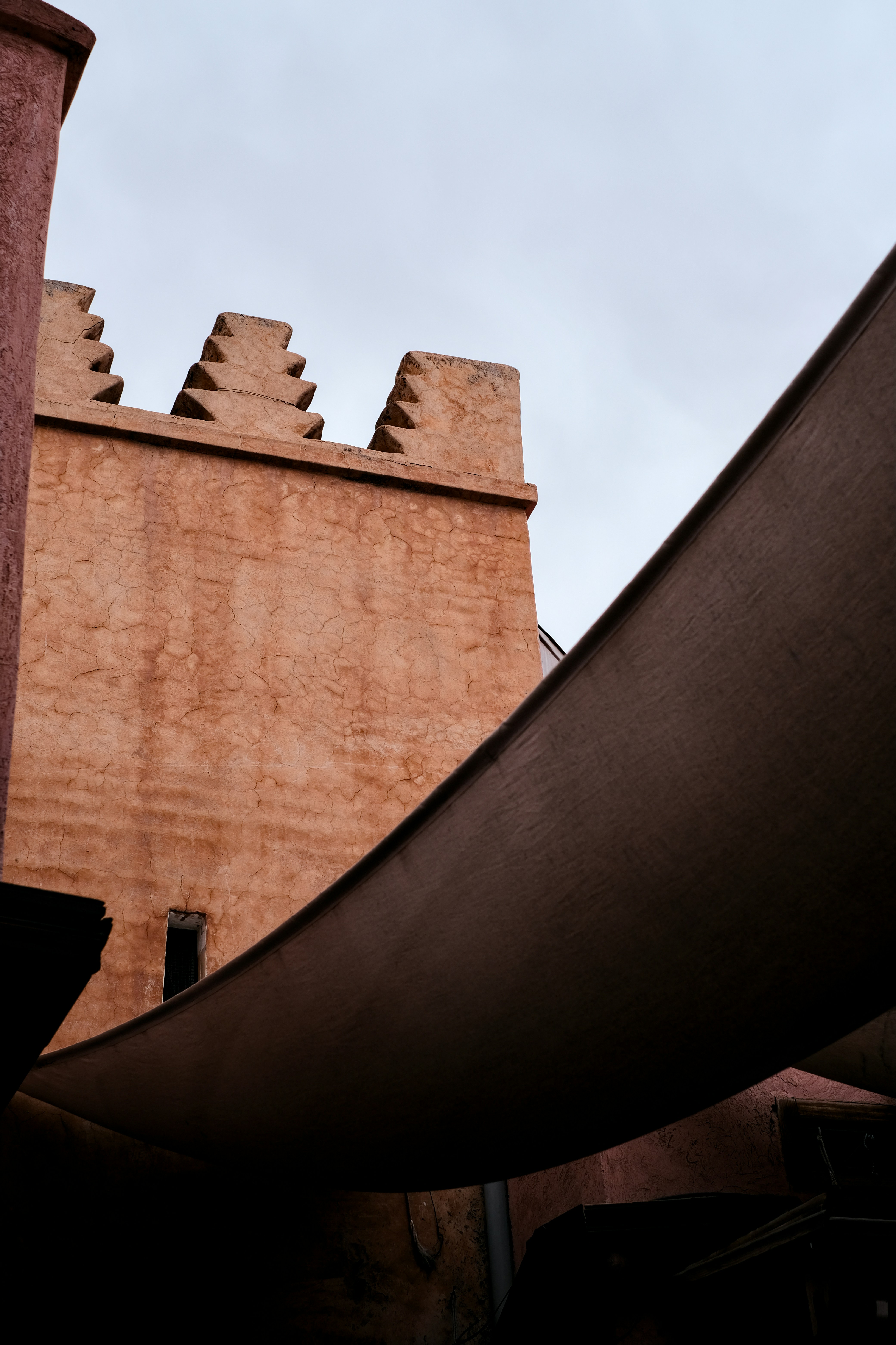 Warm-toned adobe building with distinctive stepped battlements framed by a sweeping canvas awning under a cloudy sky.