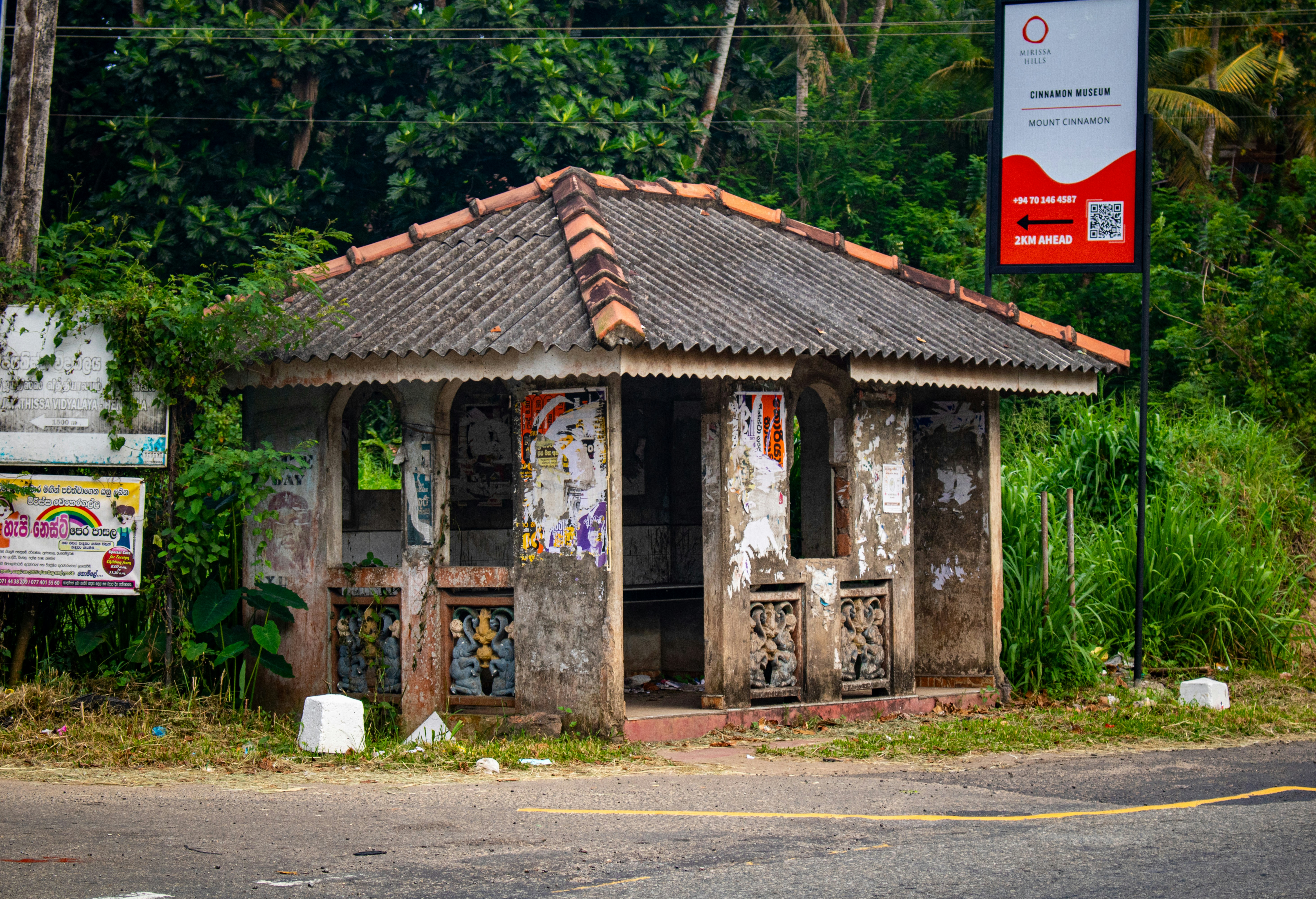 Old roadside shelter with peeling paint and posters, situated beside a lush, green highway.
