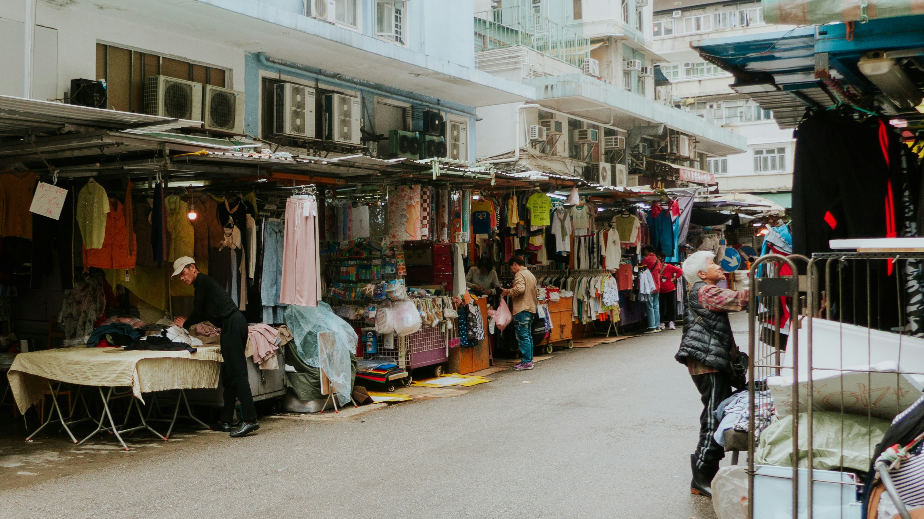 Busy market street in Hong Kong with stalls displaying clothing and goods under colorful awnings.