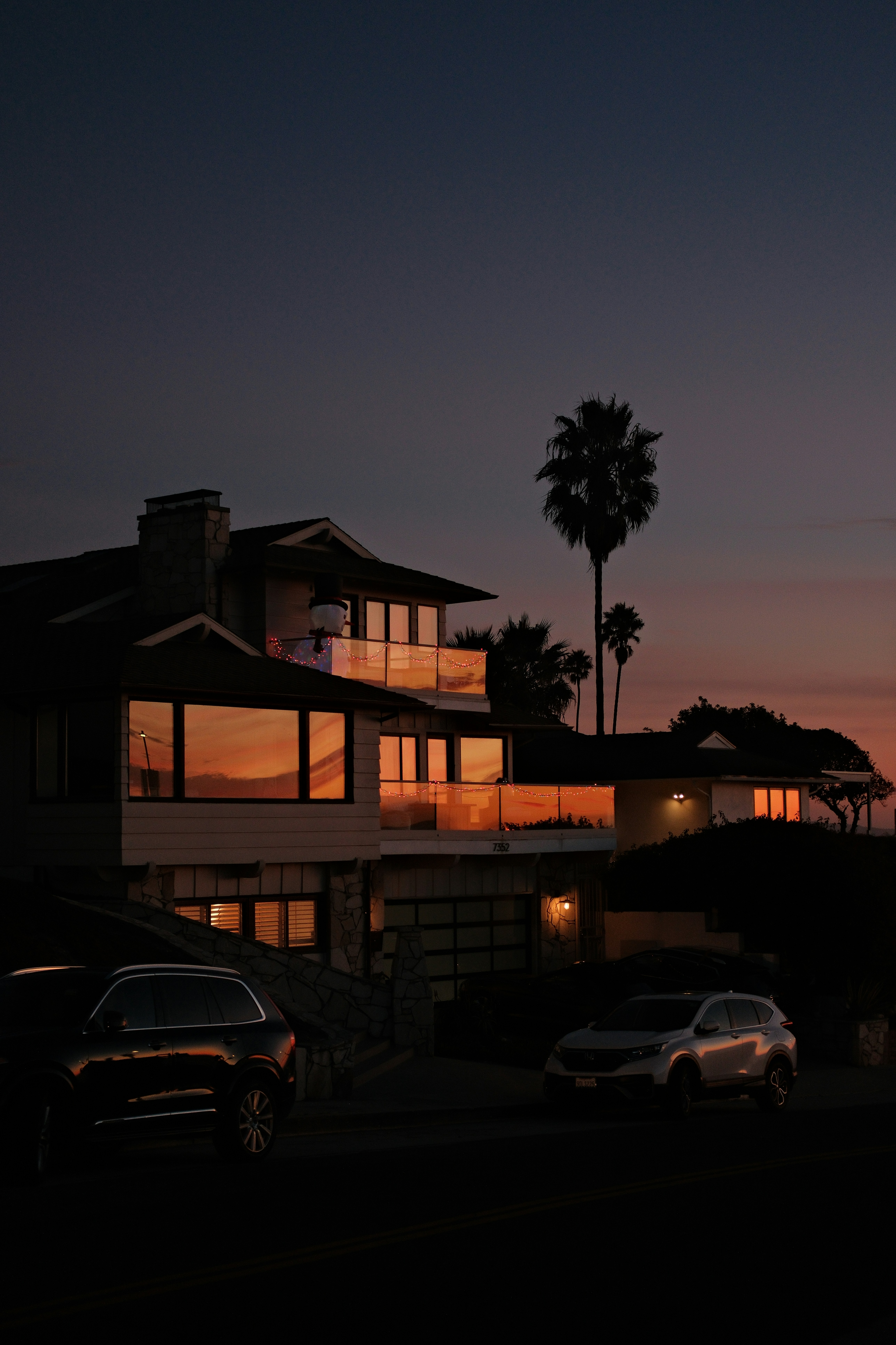 Modern house with large windows reflecting a vibrant sunset, framed by silhouetted palm trees.