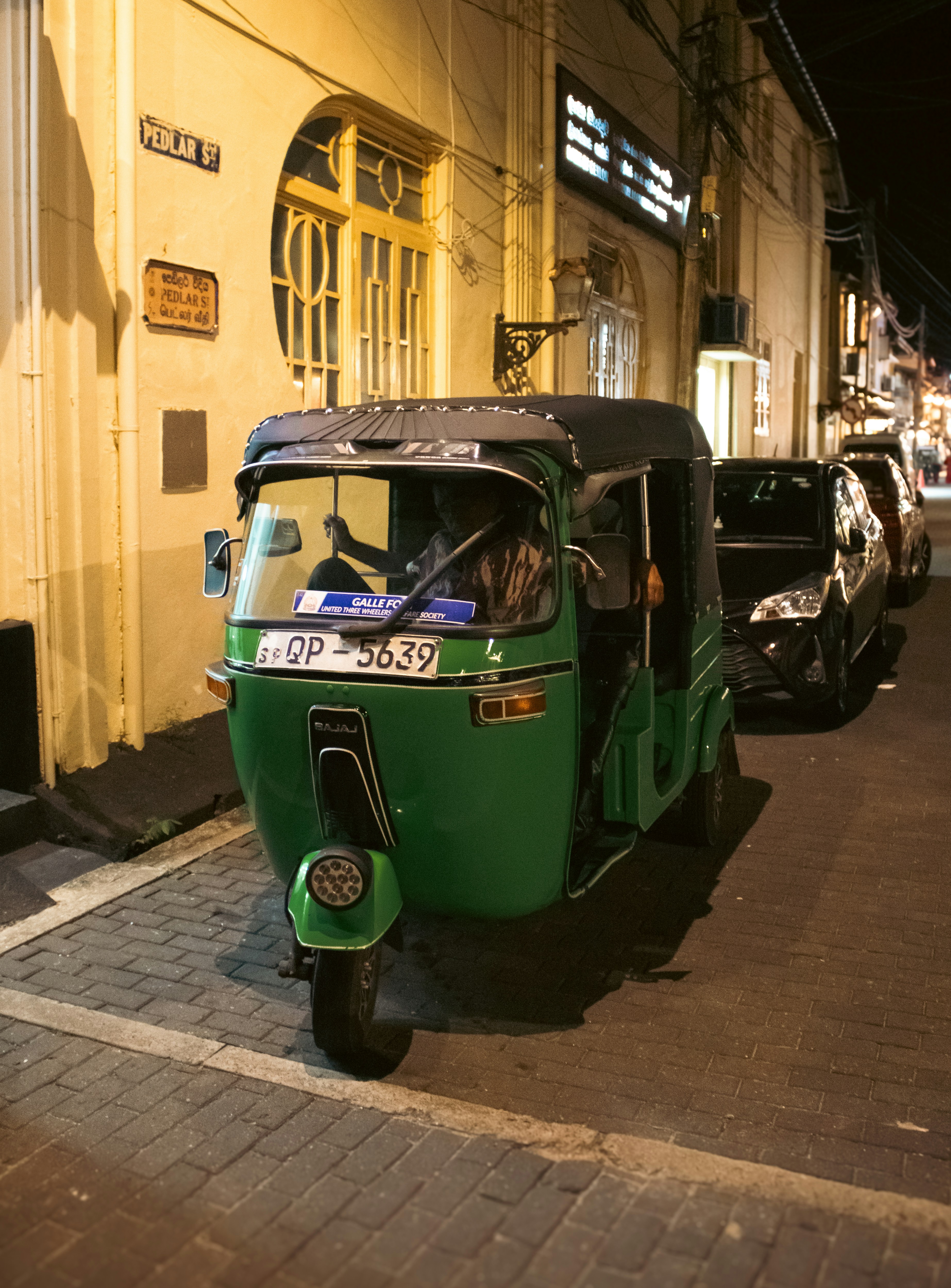Amber-lit cobblestone street cradles a green tuk-tuk