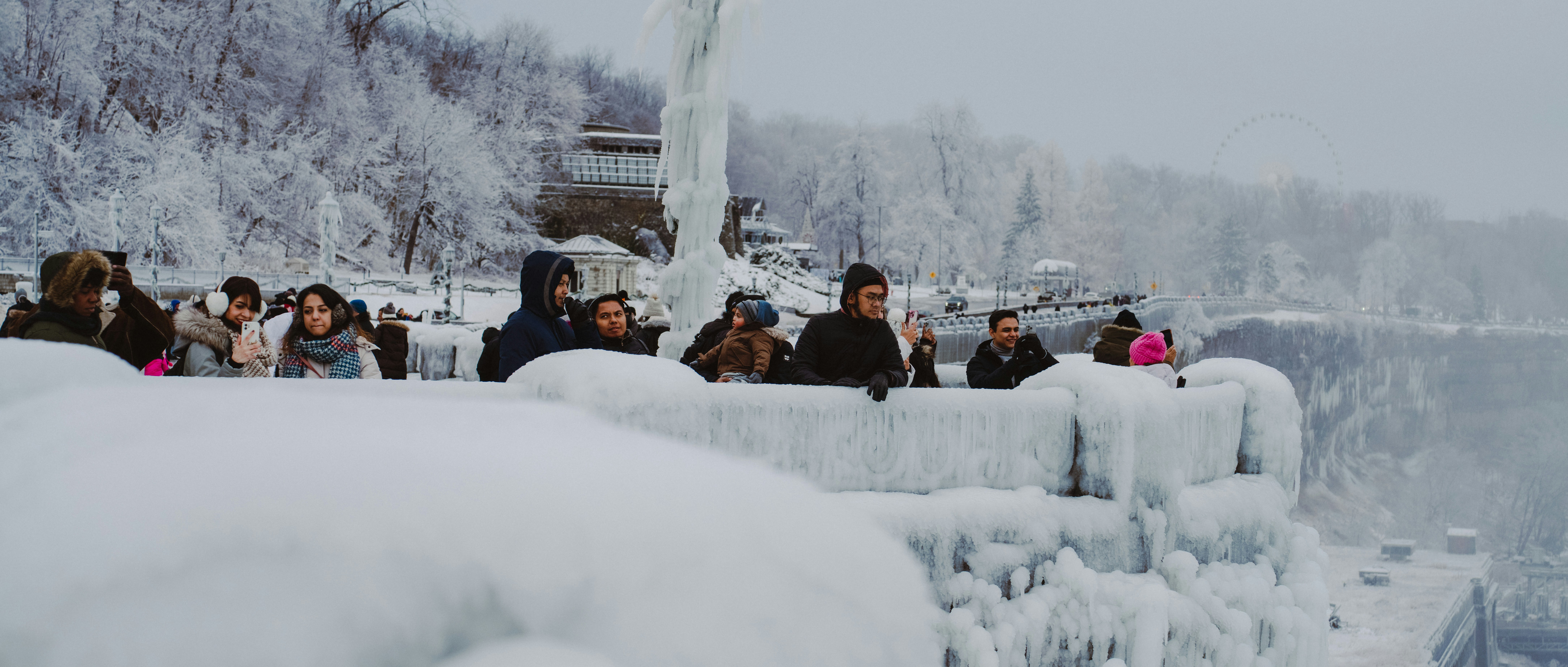 Visitors bundled up against the cold, taking in the stunning frozen scenery at Niagara Falls. Thick layers of ice and snow cover the railings and trees, transforming the landscape into a true winter wonderland. The crisp air, bundled figures, and frosty surroundings capture the magic of winter in Canada, with the falls in the background and a peaceful, frozen beauty all around. | People are viewing a snowy waterfall.