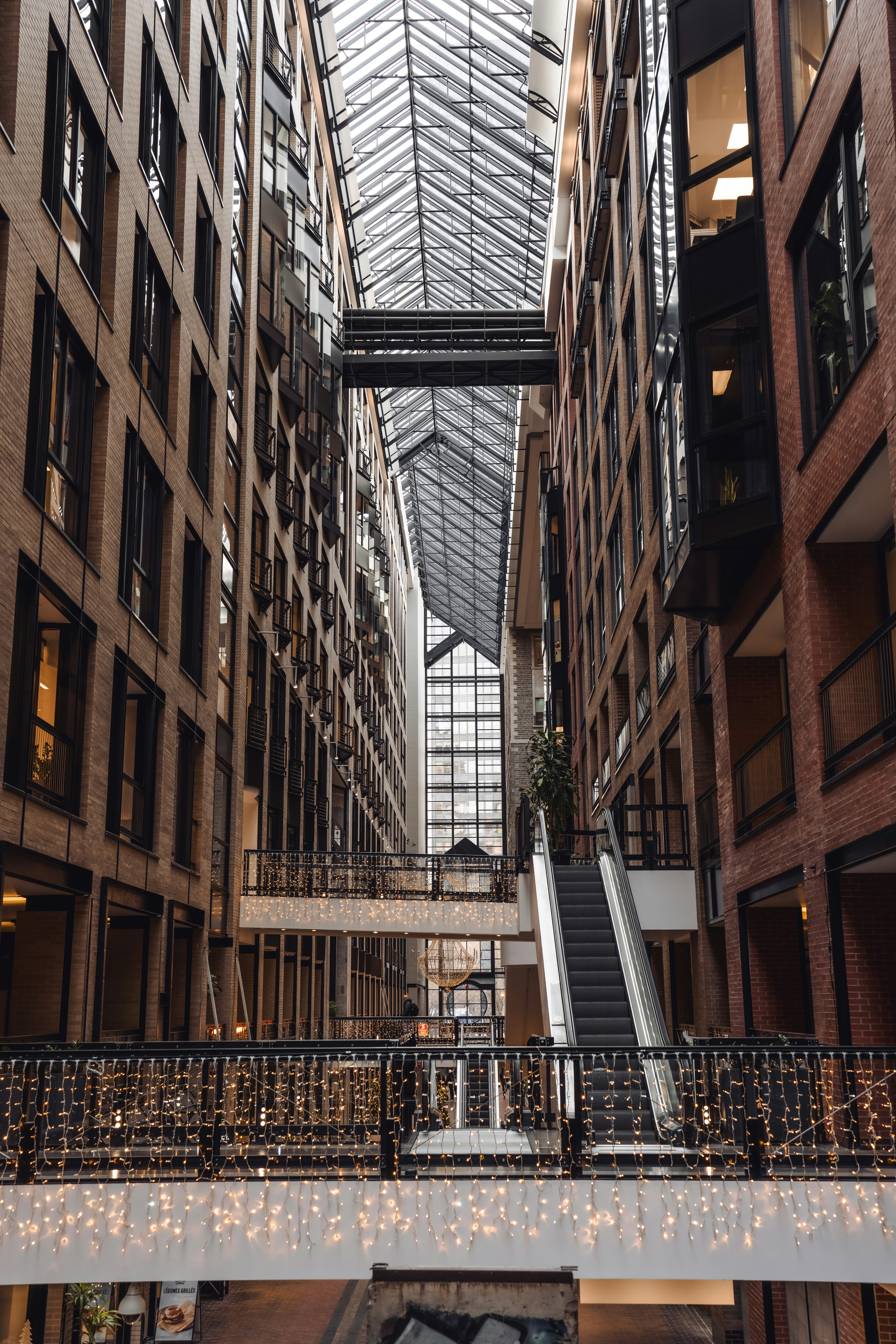 A stylish urban atrium in Montreal, featuring symmetrical brick buildings, large windows, and a glass ceiling letting in natural light. Festive string lights and a central escalator add warmth and depth, blending contemporary architecture with cozy, inviting details in this city apartment and office space.