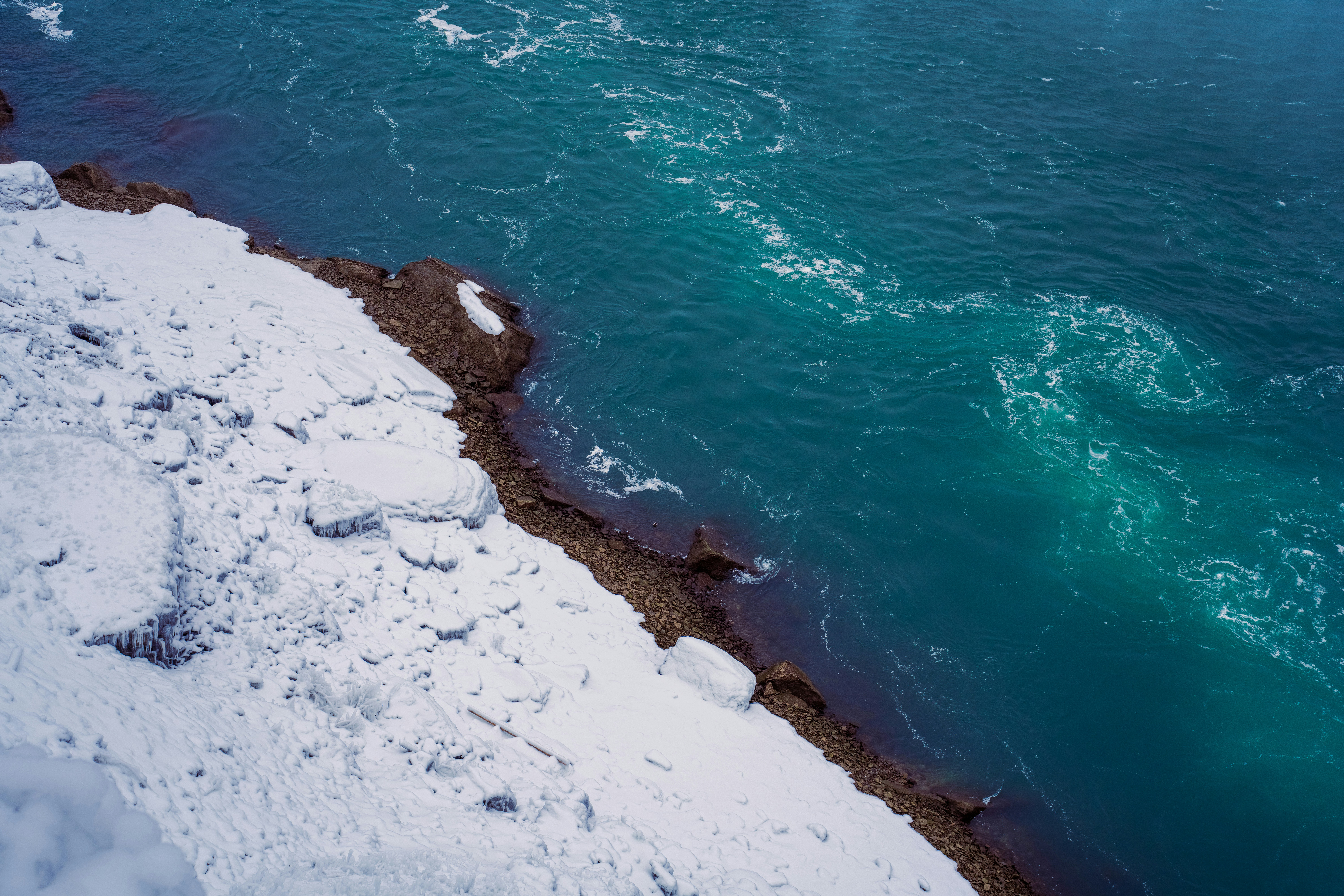 A dramatic aerial view of the icy shoreline at Niagara Falls, where crisp white snow meets the vibrant turquoise water. The rugged edge of the coast highlights the striking contrast between winter’s chill and the powerful flow of the river, capturing the raw beauty of the Canadian landscape from above.