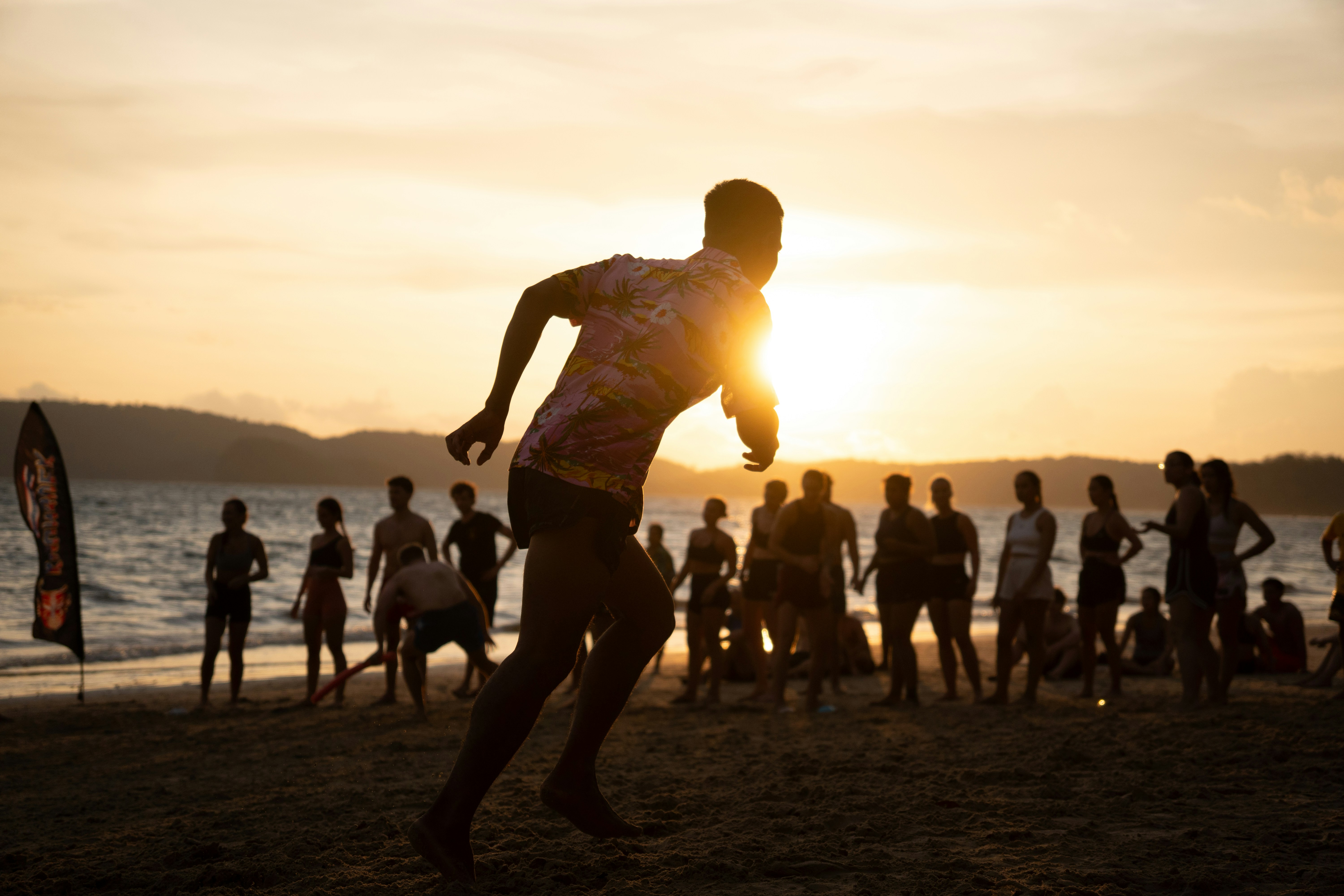 Silhouetted soccer player in action on a beach at sunset with a crowd in the background.