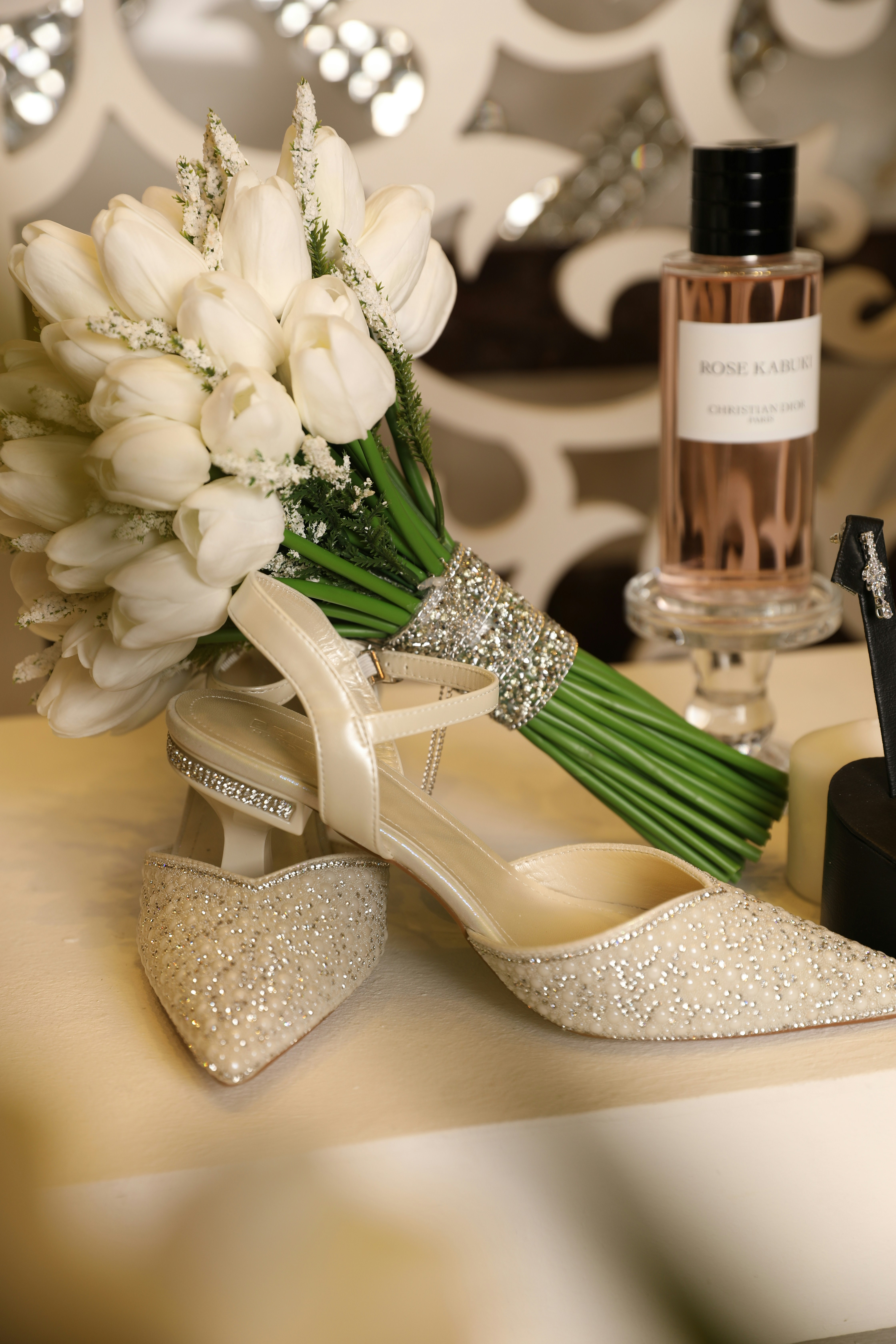 A beautifully arranged bouquet of white tulips rests on a pair of sparkling bridal shoes, accompanied by a bottle of perfume. The scene captures the essence of wedding elegance.