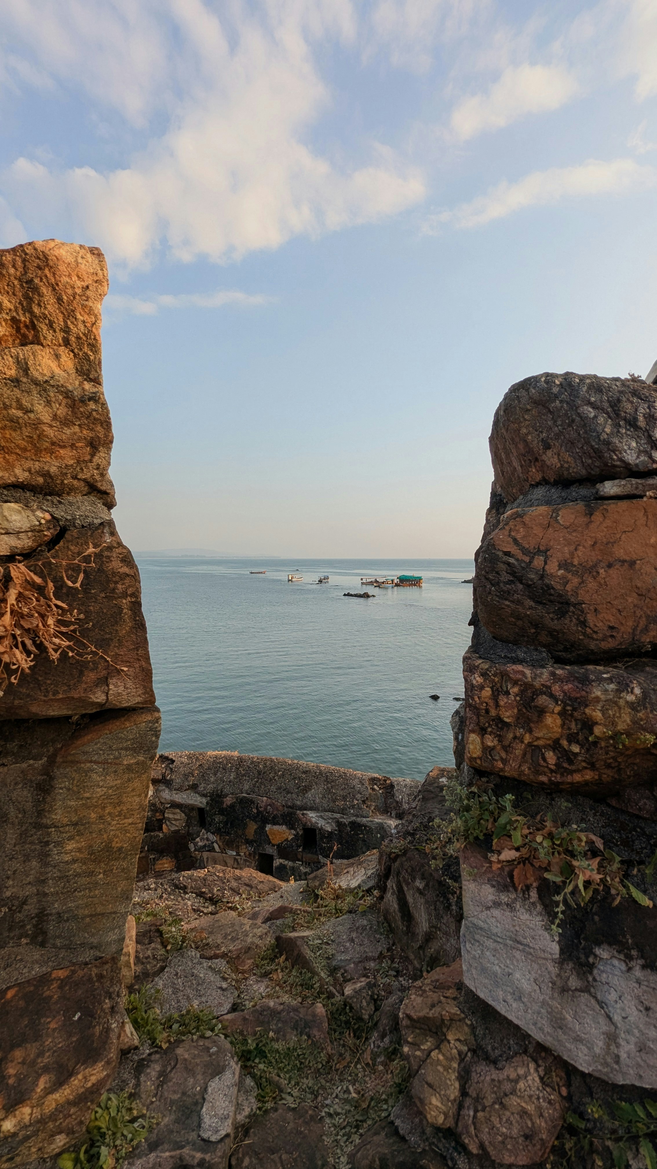 Sunlit sea view framed by ancient stone walls, with small boats dotting the calm water beneath a clear sky.
