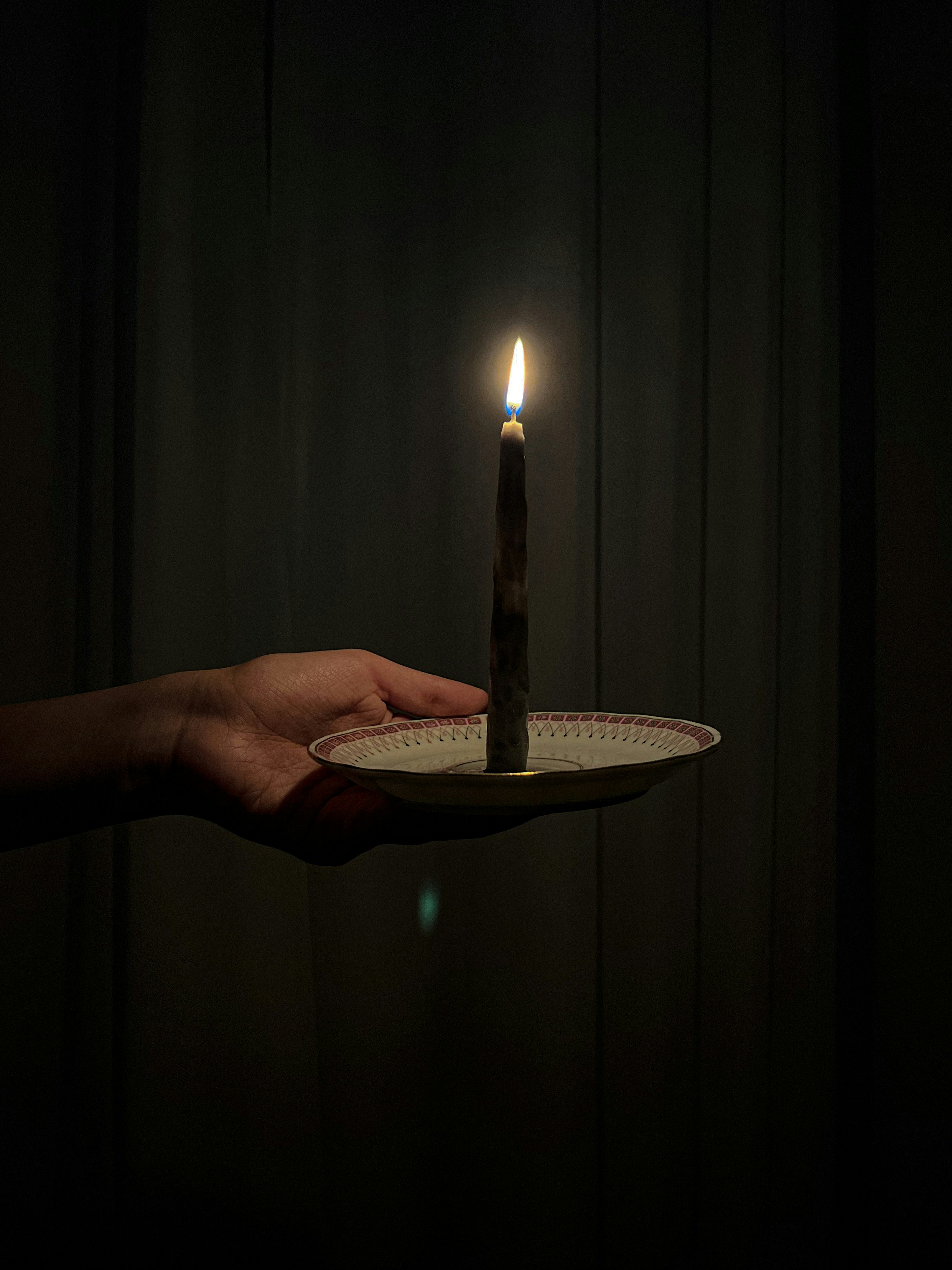 A hand holds a candle atop a decorative plate, illuminating the dim surroundings with a soft glow.