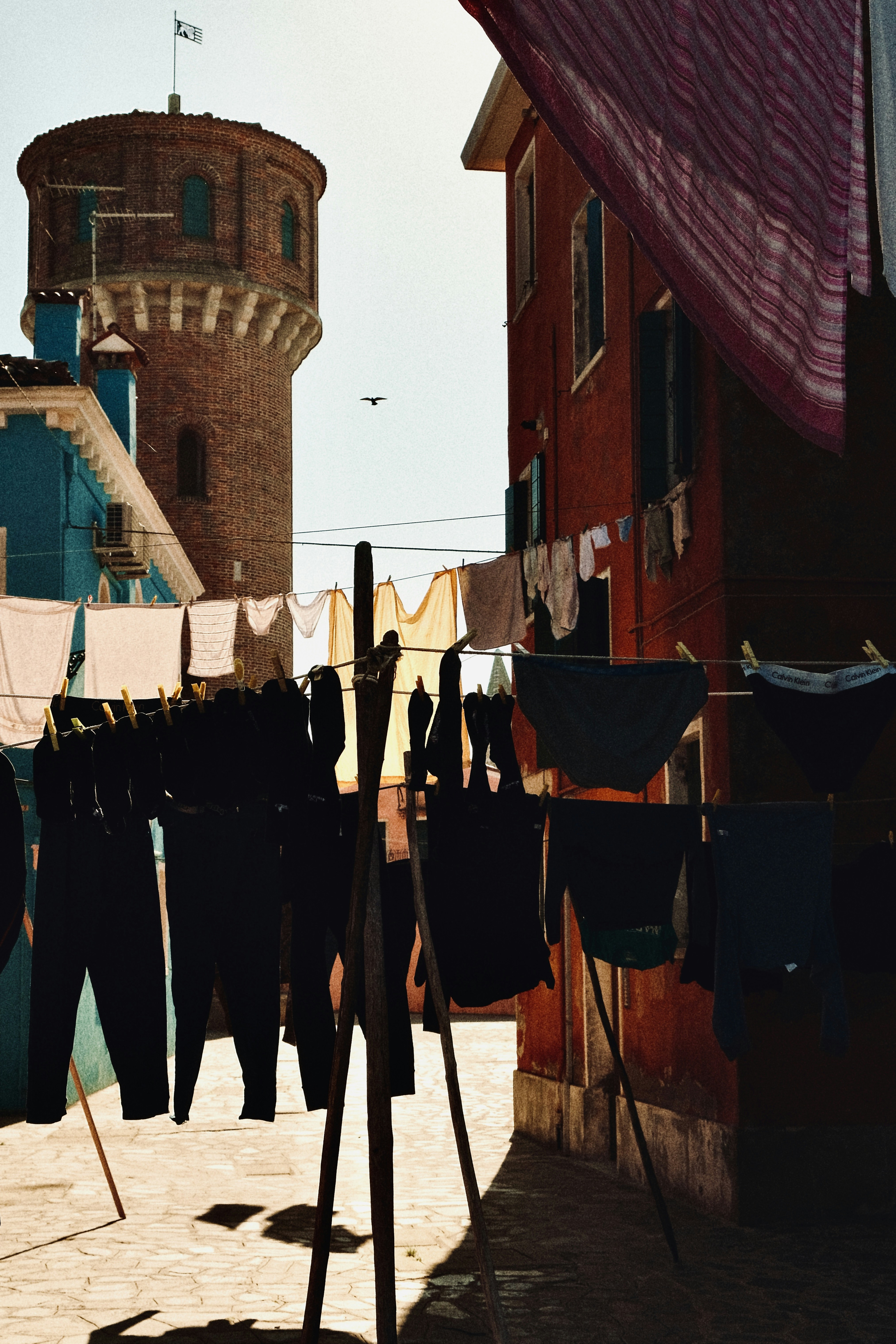 Colorful clotheslines cast dramatic shadows in a narrow alley beneath a bright tower in Burano, Italy.