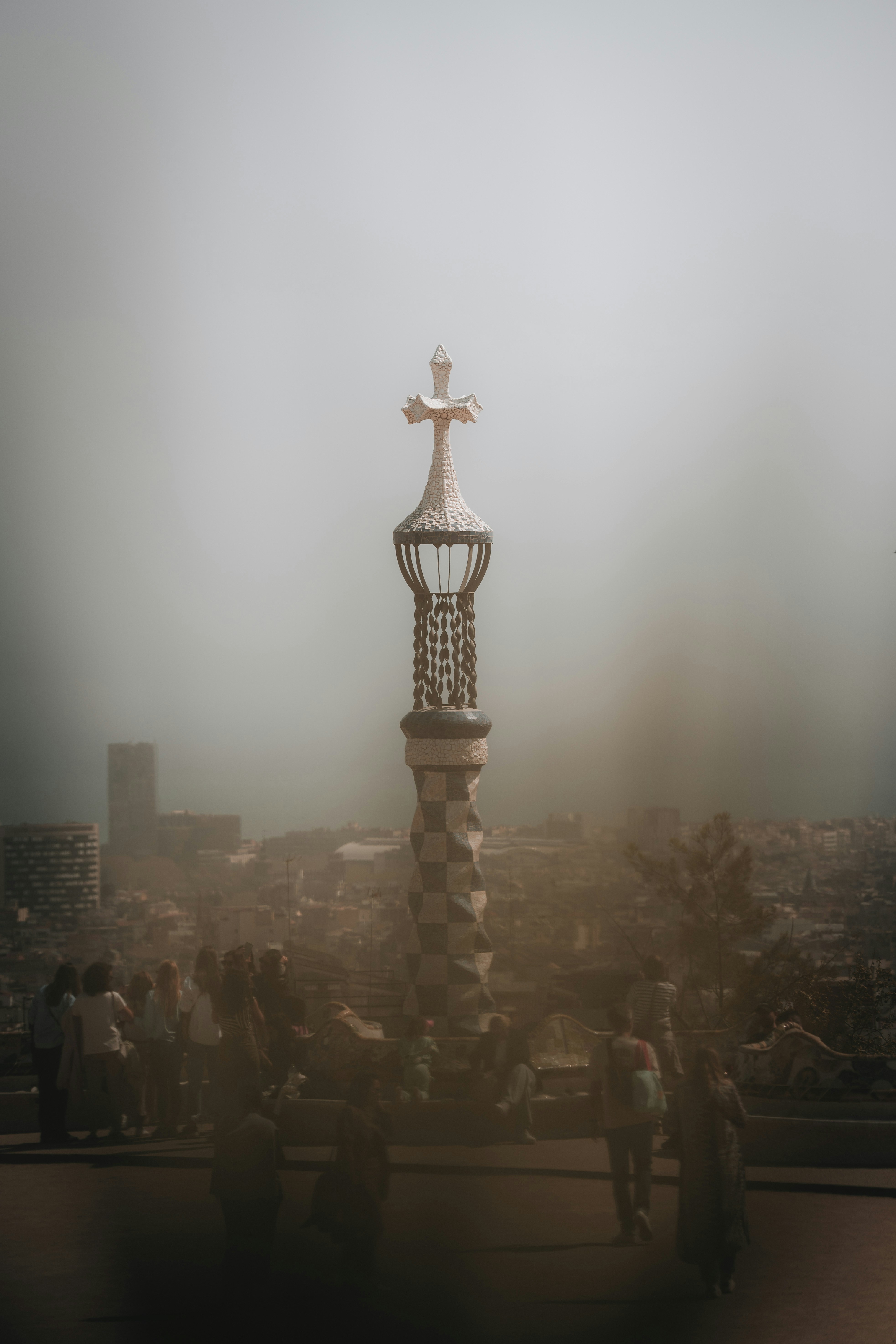 Colorful architectural tower adorned with a cross stands prominently over a bustling crowd in a cityscape. The hazy background adds a dreamlike quality.