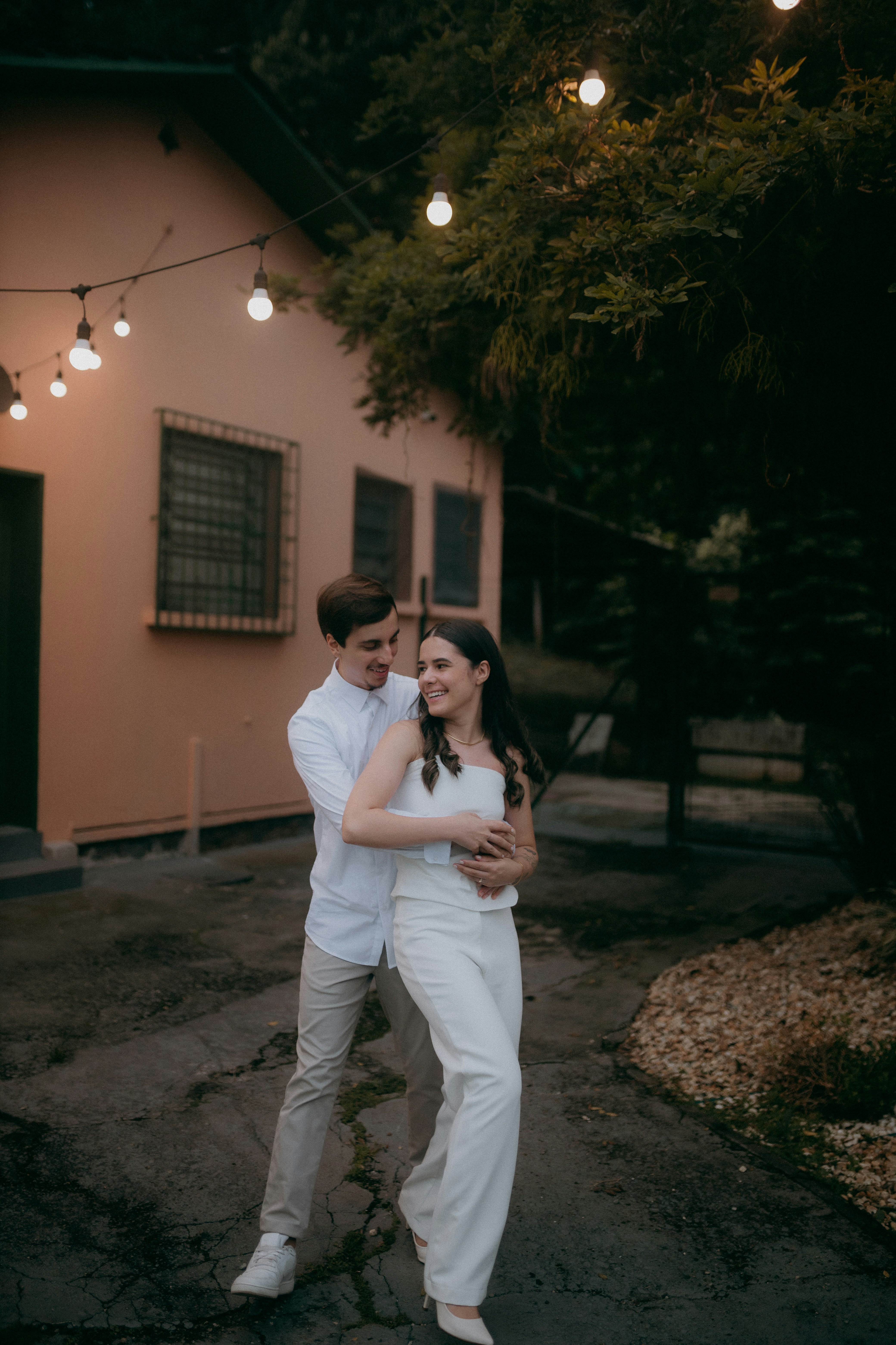 Couple embracing on a dimly lit patio with string lights overhead and a rustic building in the background.