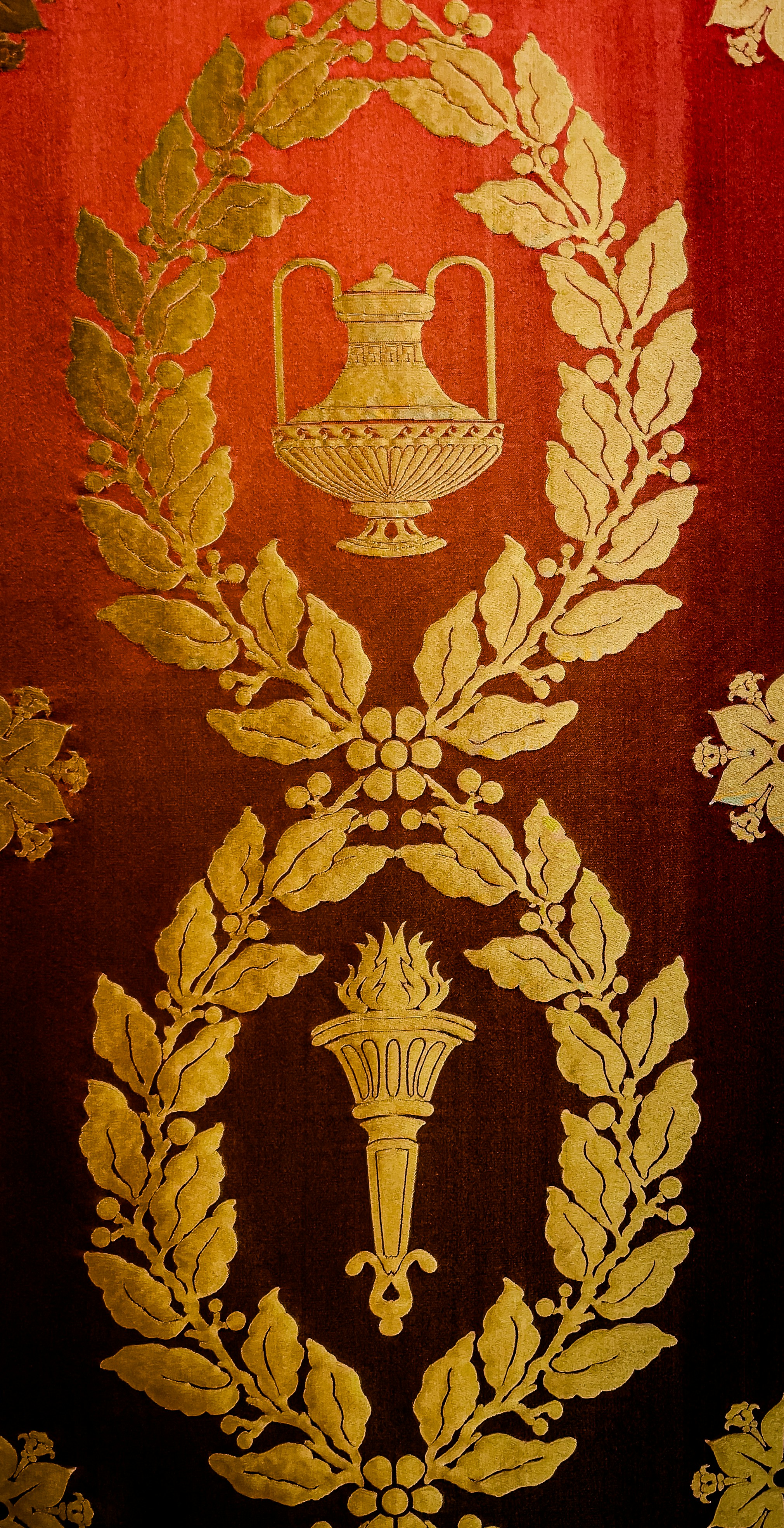 Intricate golden motifs of a vase and a torch surrounded by lush leaves, set against a rich red backdrop.