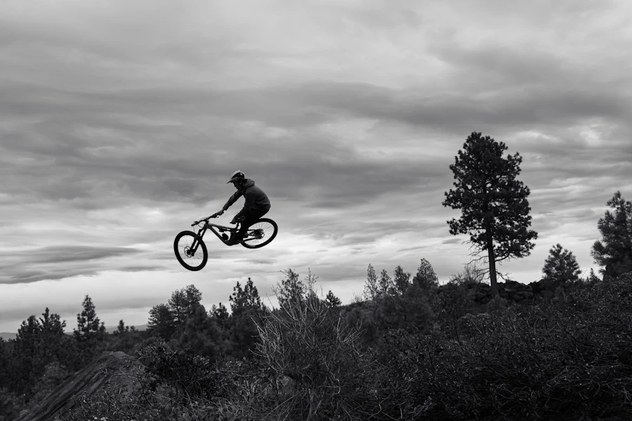 Mountain biker launching a jump over shrub-covered ridge, silhouetted against dramatic cloud cover.