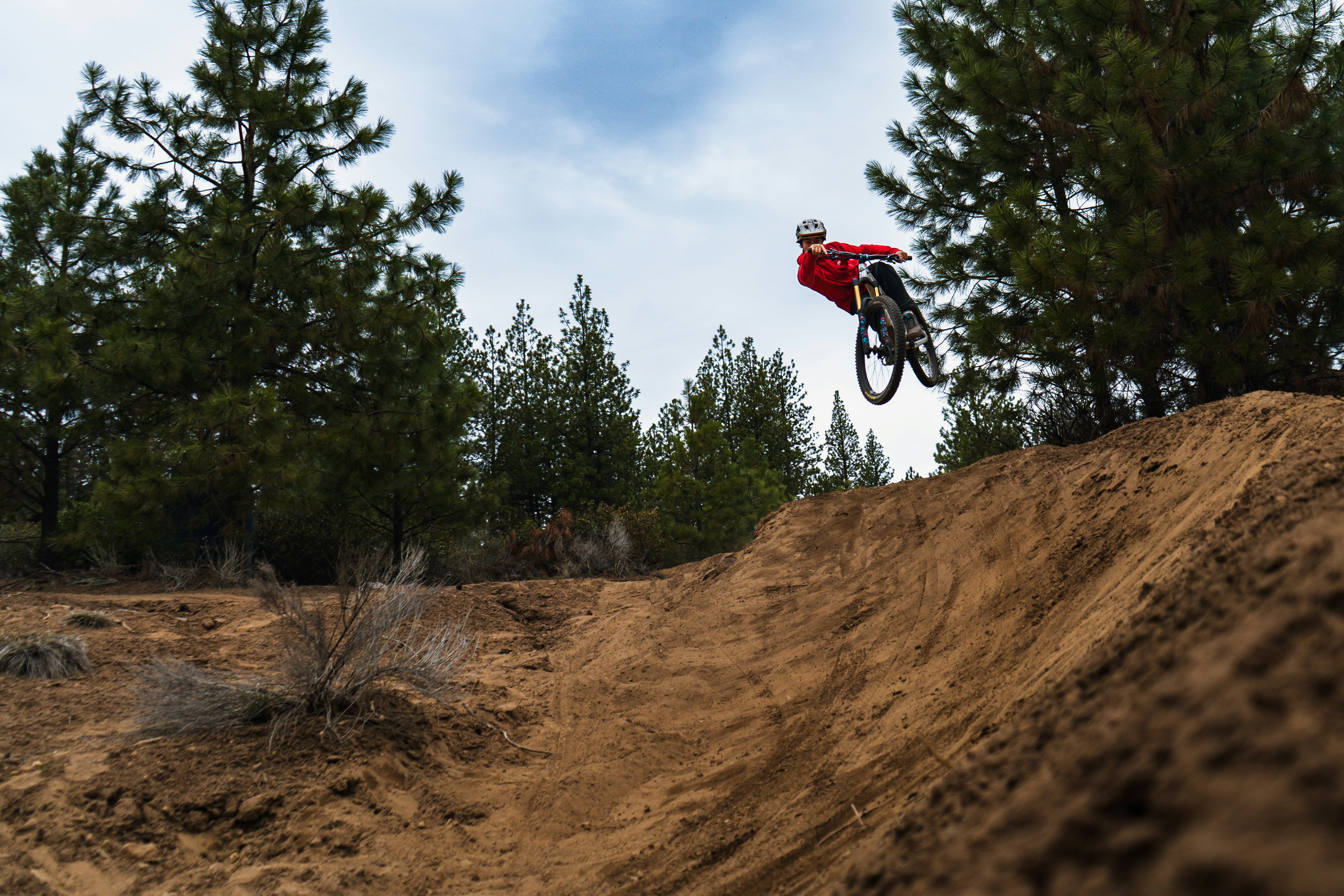 Cyclist in a red jersey launches off a dirt ramp, airborne above a forested trail with tall pines framing the scene.