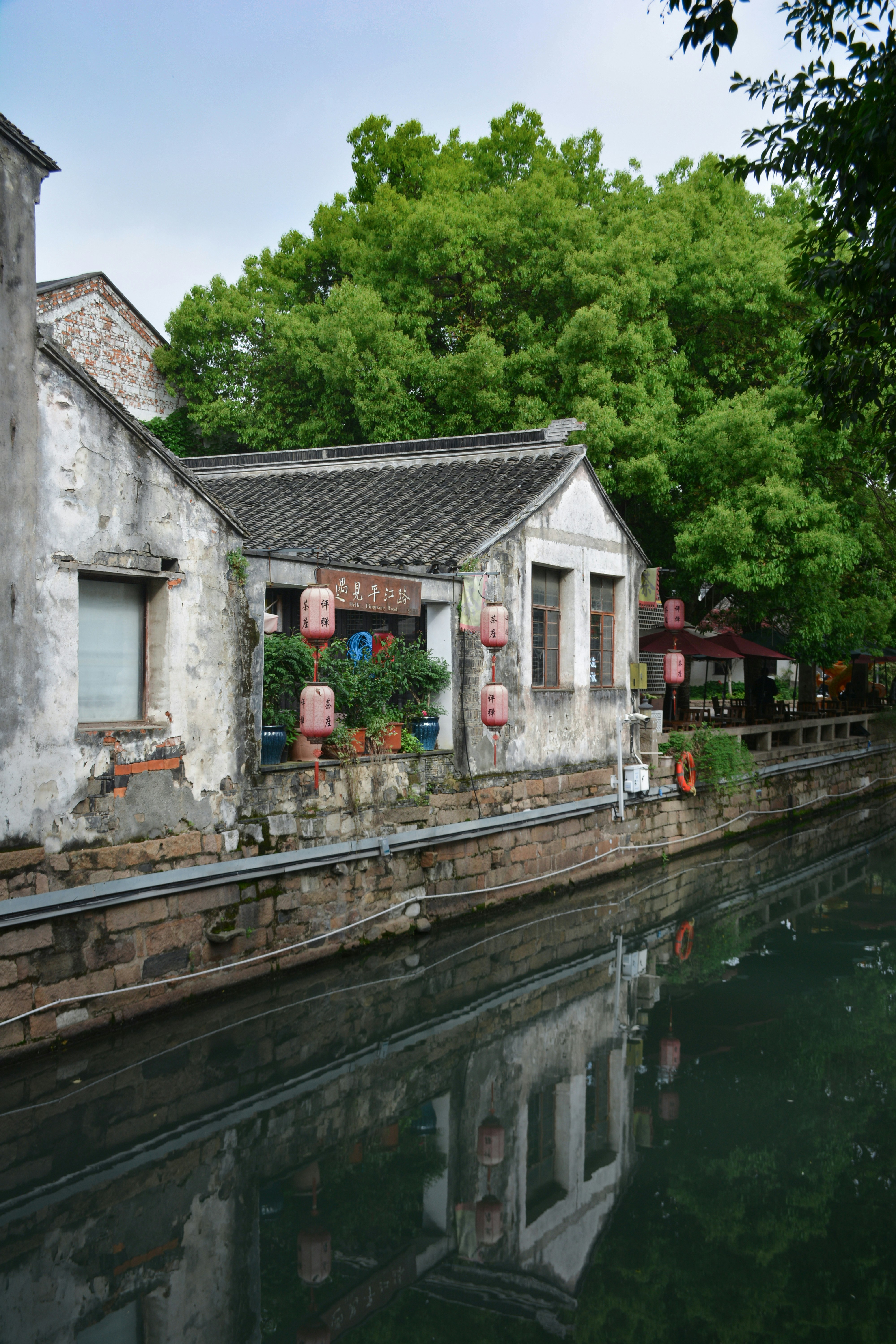 Traditional canal-side building with red lanterns and lush green trees reflecting in the water.