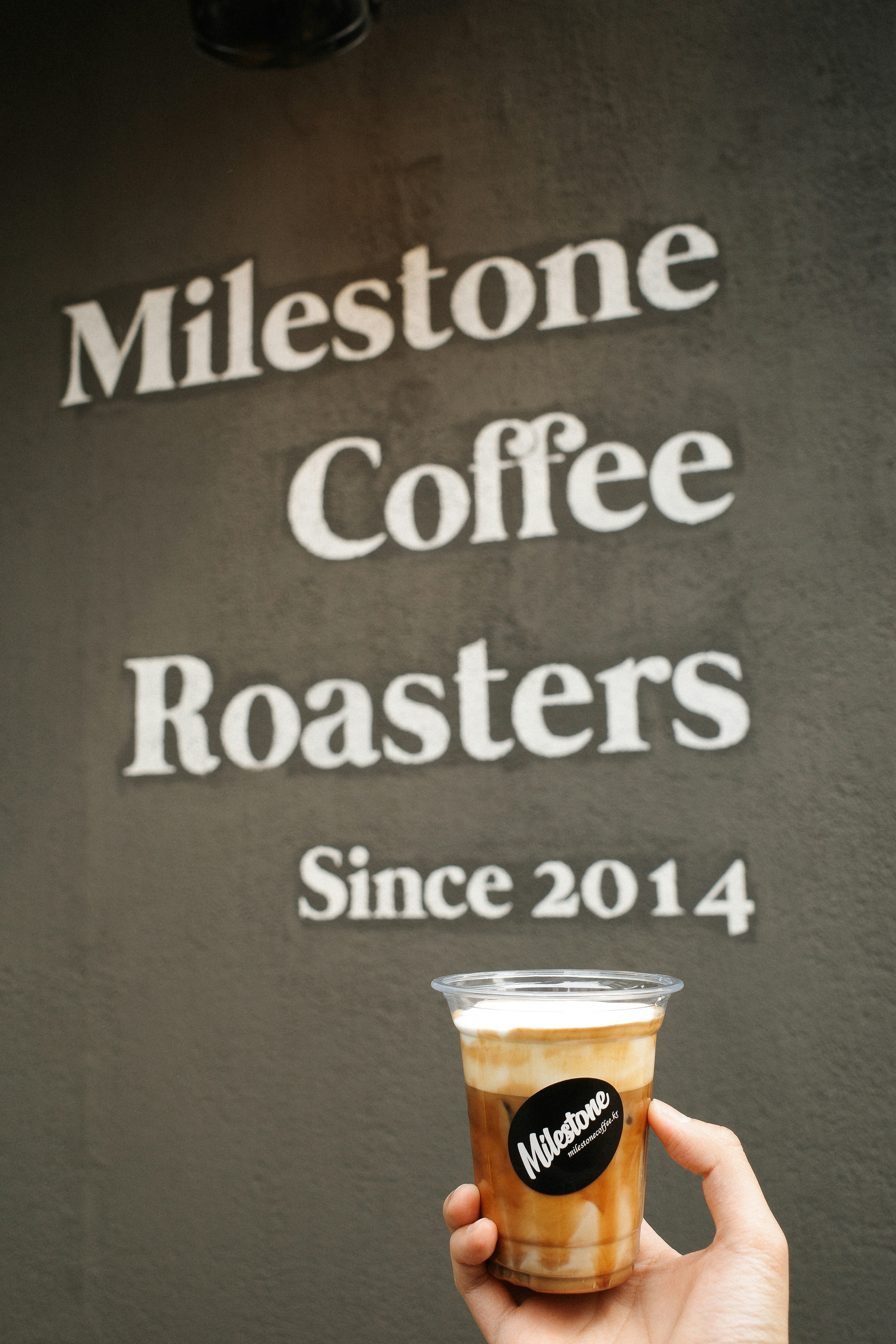 Milestone Latte Portrait: Handheld Cup Before the Roasters Wall