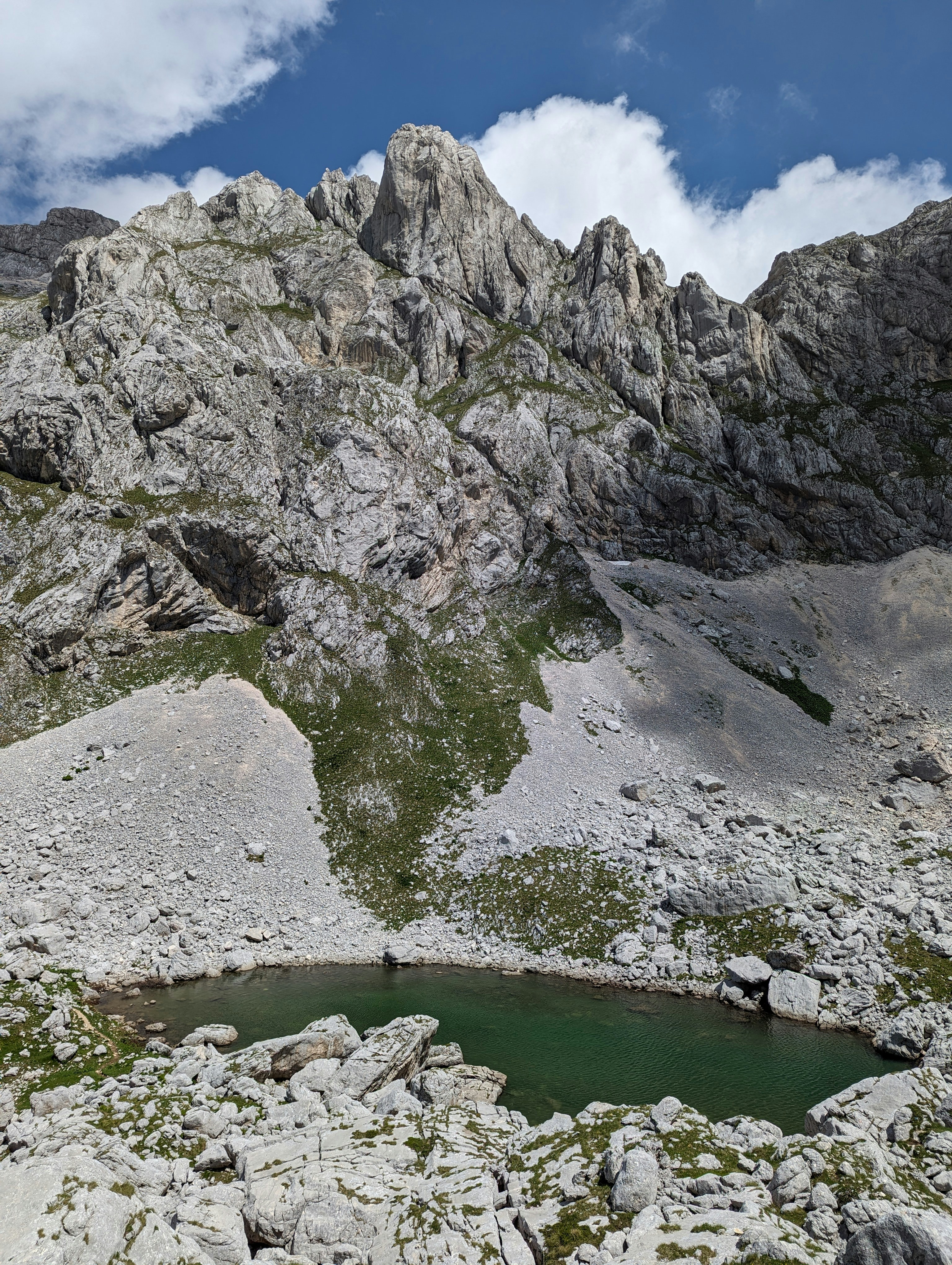 Rocky mountain landscape with a small green pond nestled at the base under a partly cloudy sky.