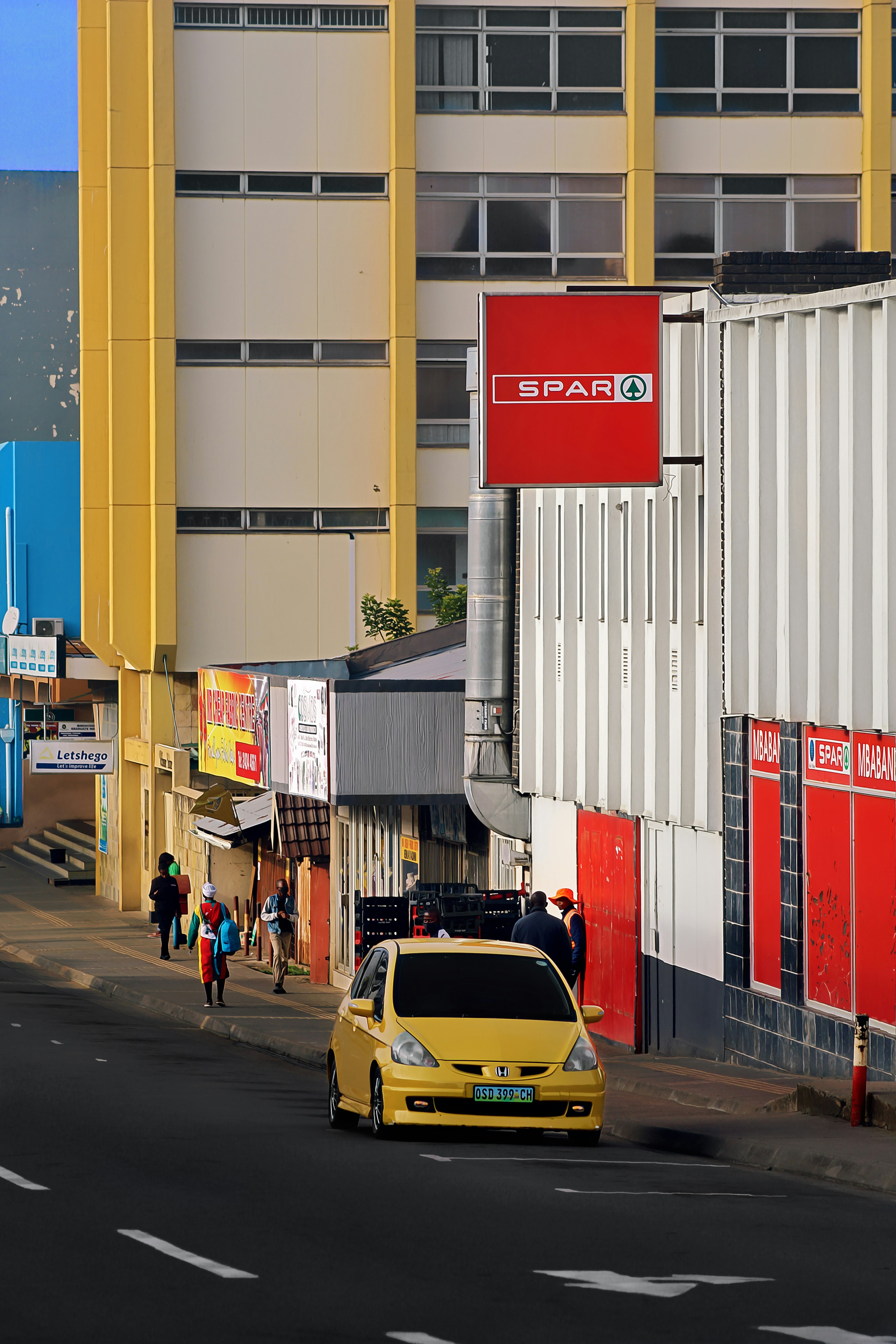 A vibrant yellow car parked on a city street, with shops and pedestrians in the background. The scene captures the essence of urban life and commerce.
