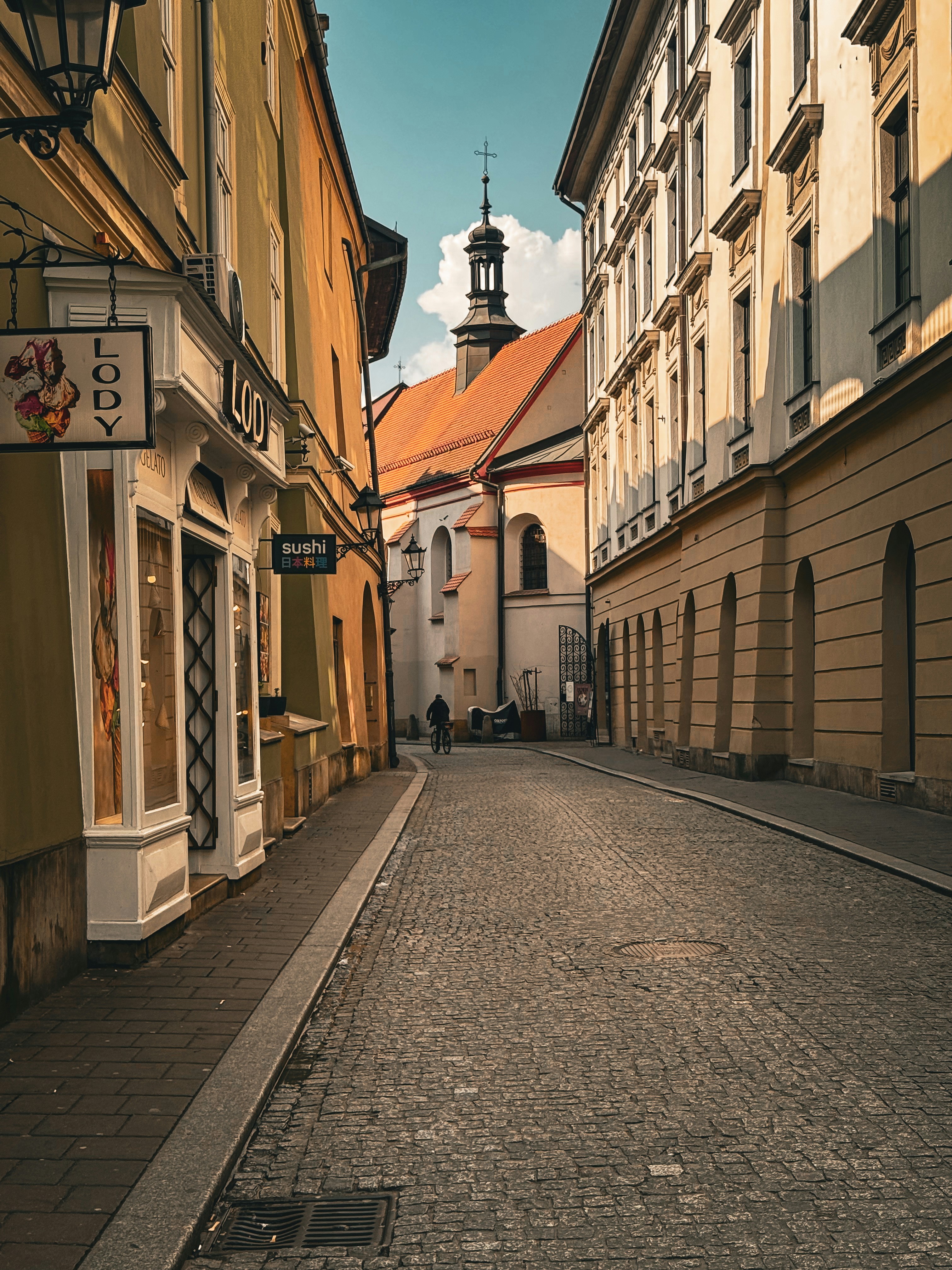 Charming alleyway lined with colorful buildings, featuring a sushi restaurant and a glimpse of a church in the distance. The cobblestone path invites exploration.