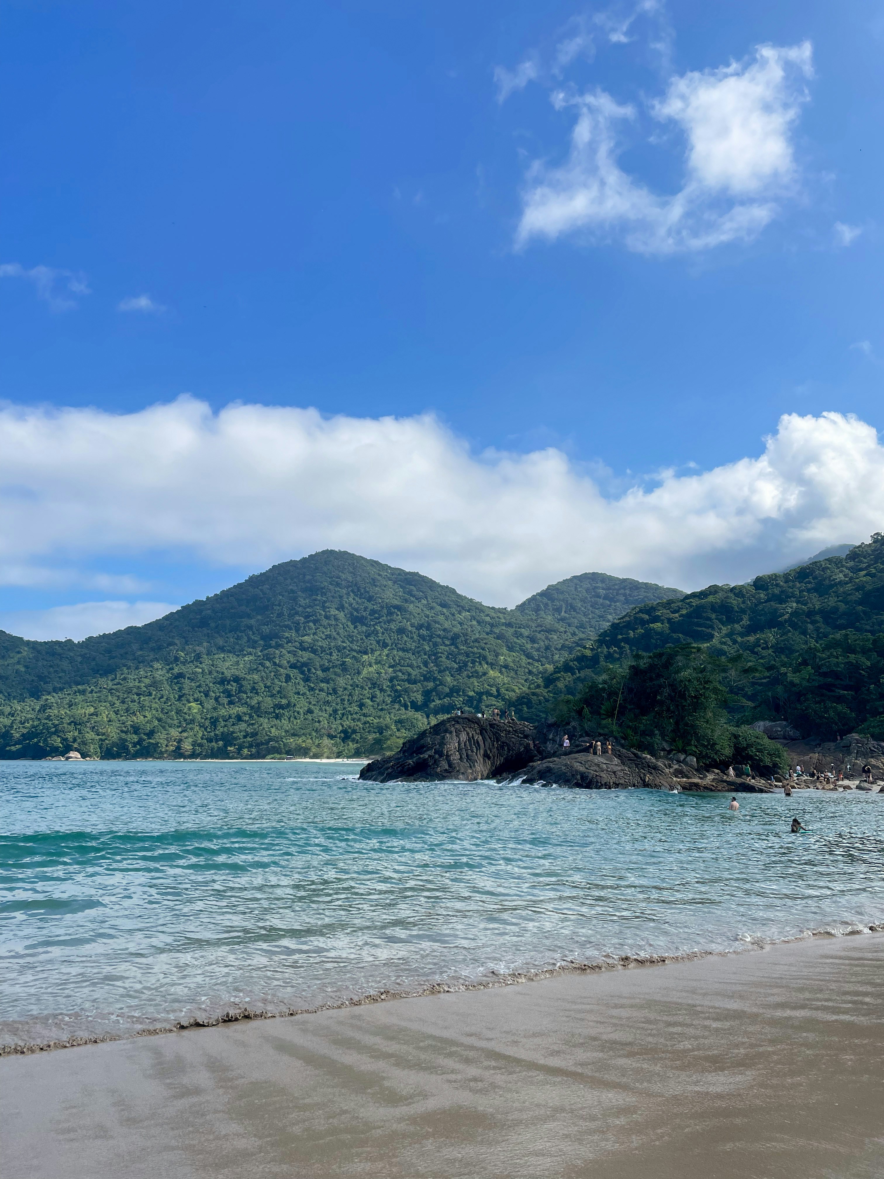 Tranquil beach scene with gentle waves lapping at the shore, framed by lush green mountains under a bright blue sky.