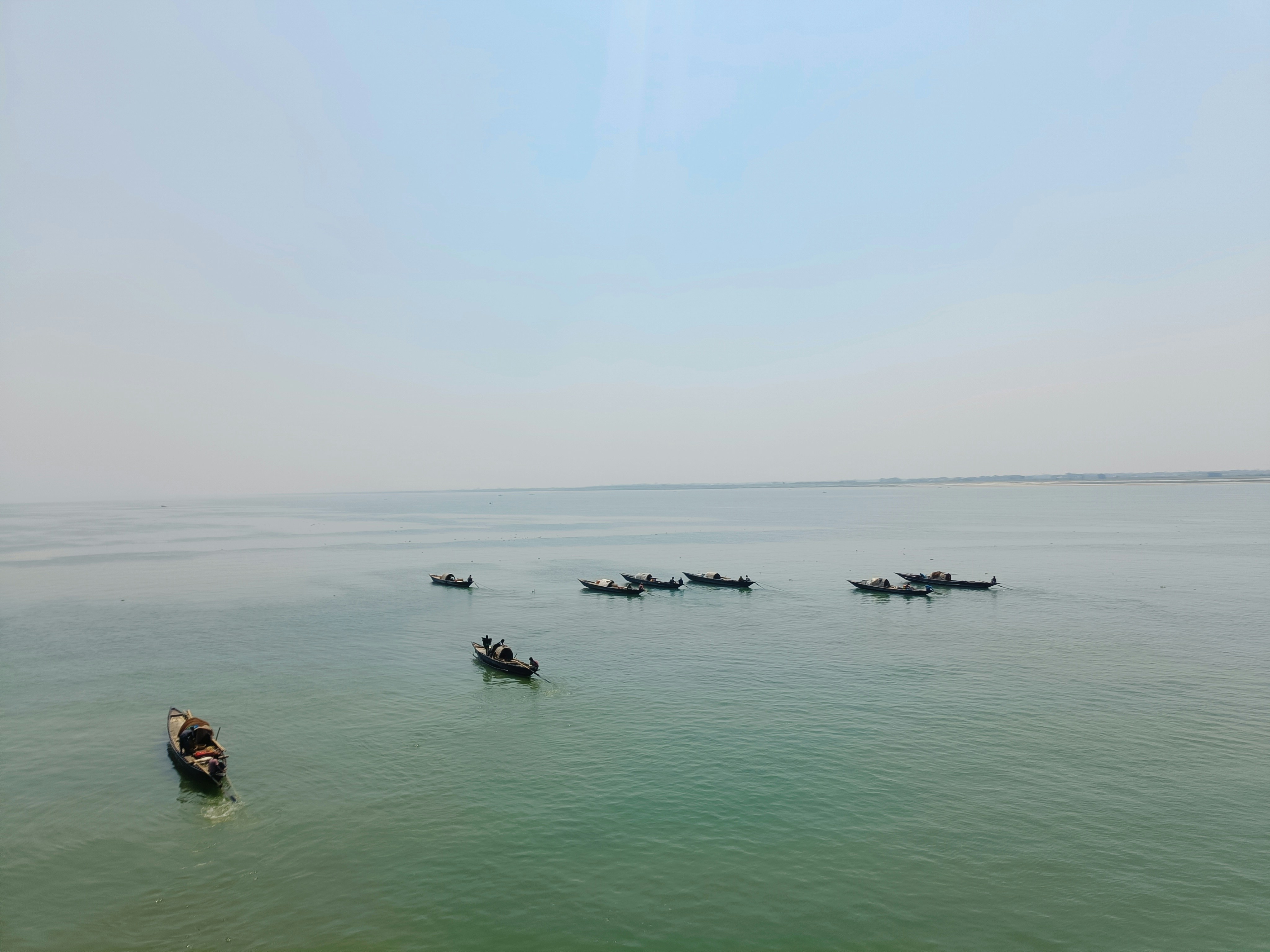 Several fishing boats glide across a calm, reflective body of water under a clear sky. The serene scene captures the essence of daily life on the water.