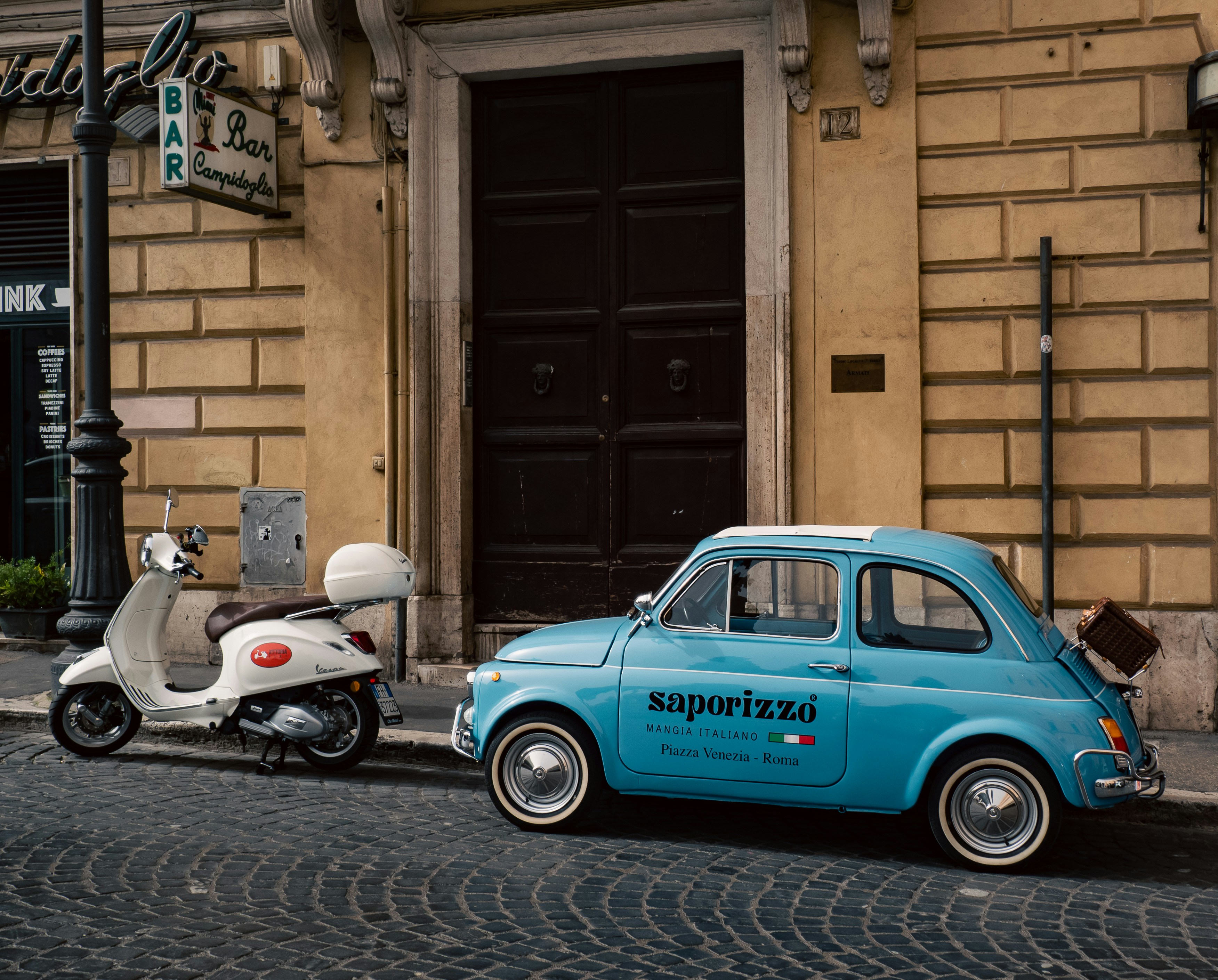 A vibrant blue vintage car parked beside a classic scooter against a historic building backdrop. The scene evokes a charming atmosphere of Italian urban life.