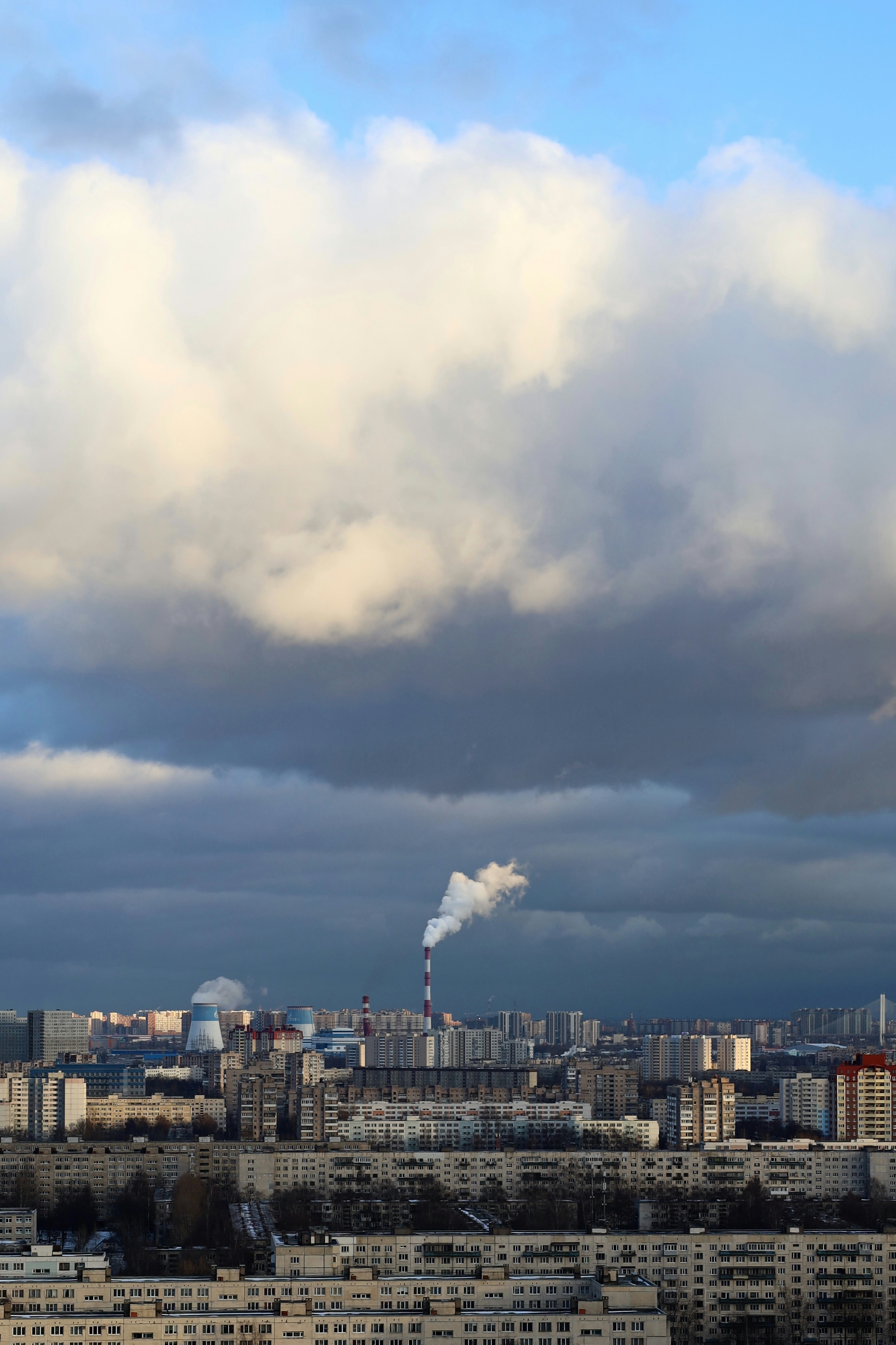 A panoramic view of a cityscape under a dramatic sky, featuring industrial smokestacks amidst residential buildings. The scene captures the contrast between urban development and atmospheric elements.