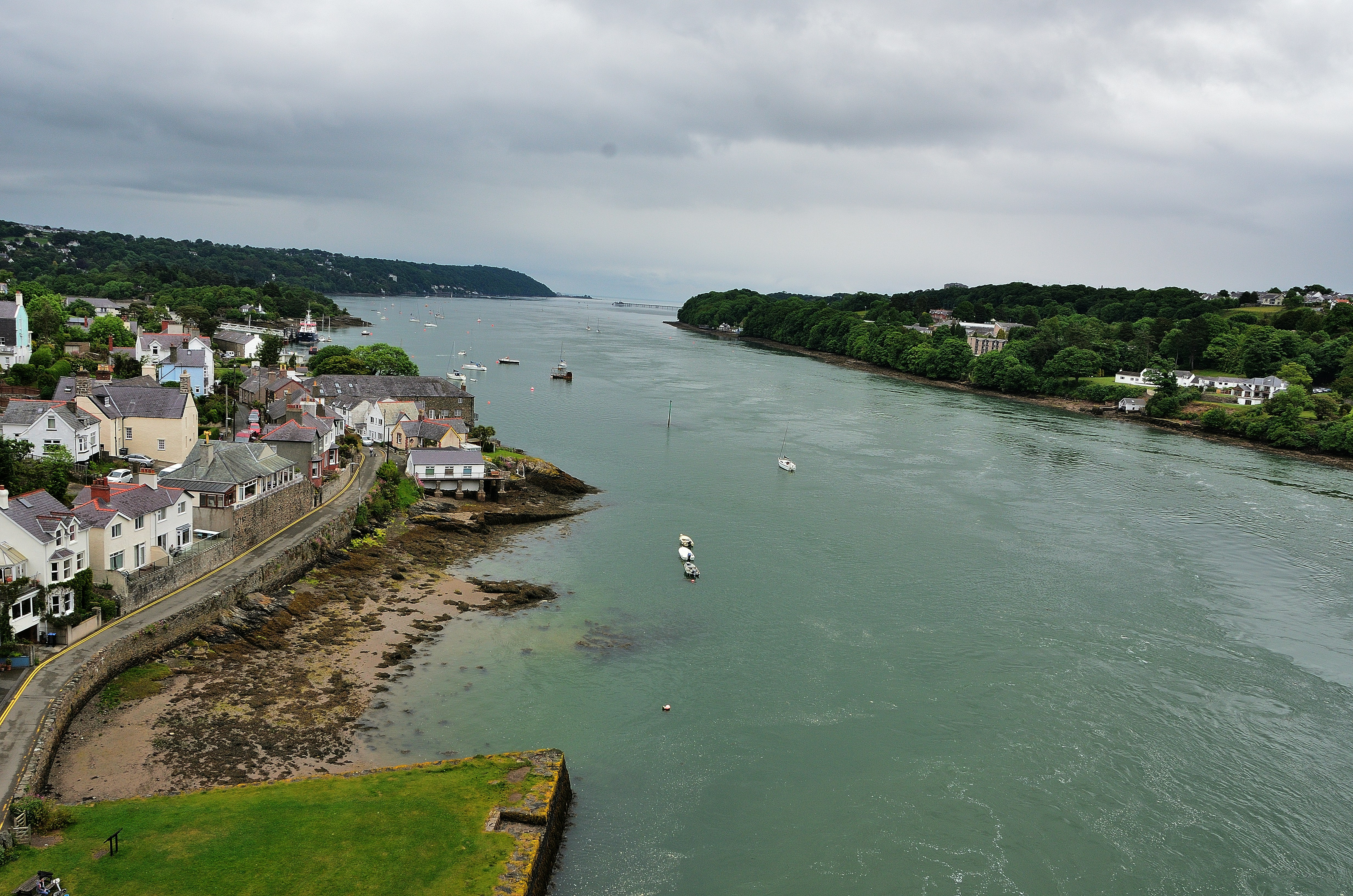 Aerial view of the Menai Strait with coastal towns and lush greenery under a cloudy sky.
