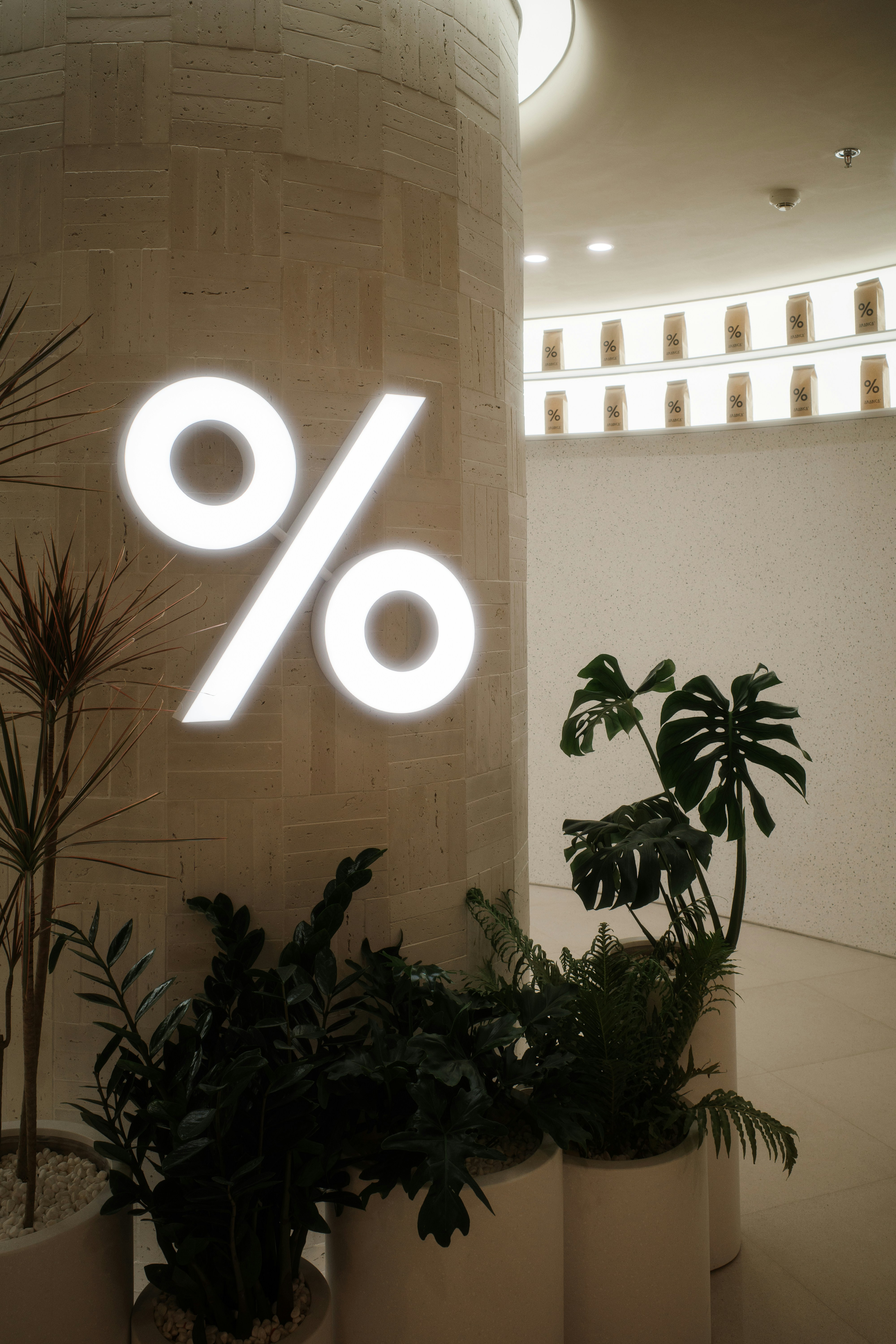 An oversized, glowing percent sign dominates a textured column in a modern interior, with lush potted plants at the base. In the background, a curved wall hosts a row of small, evenly spaced display niches.