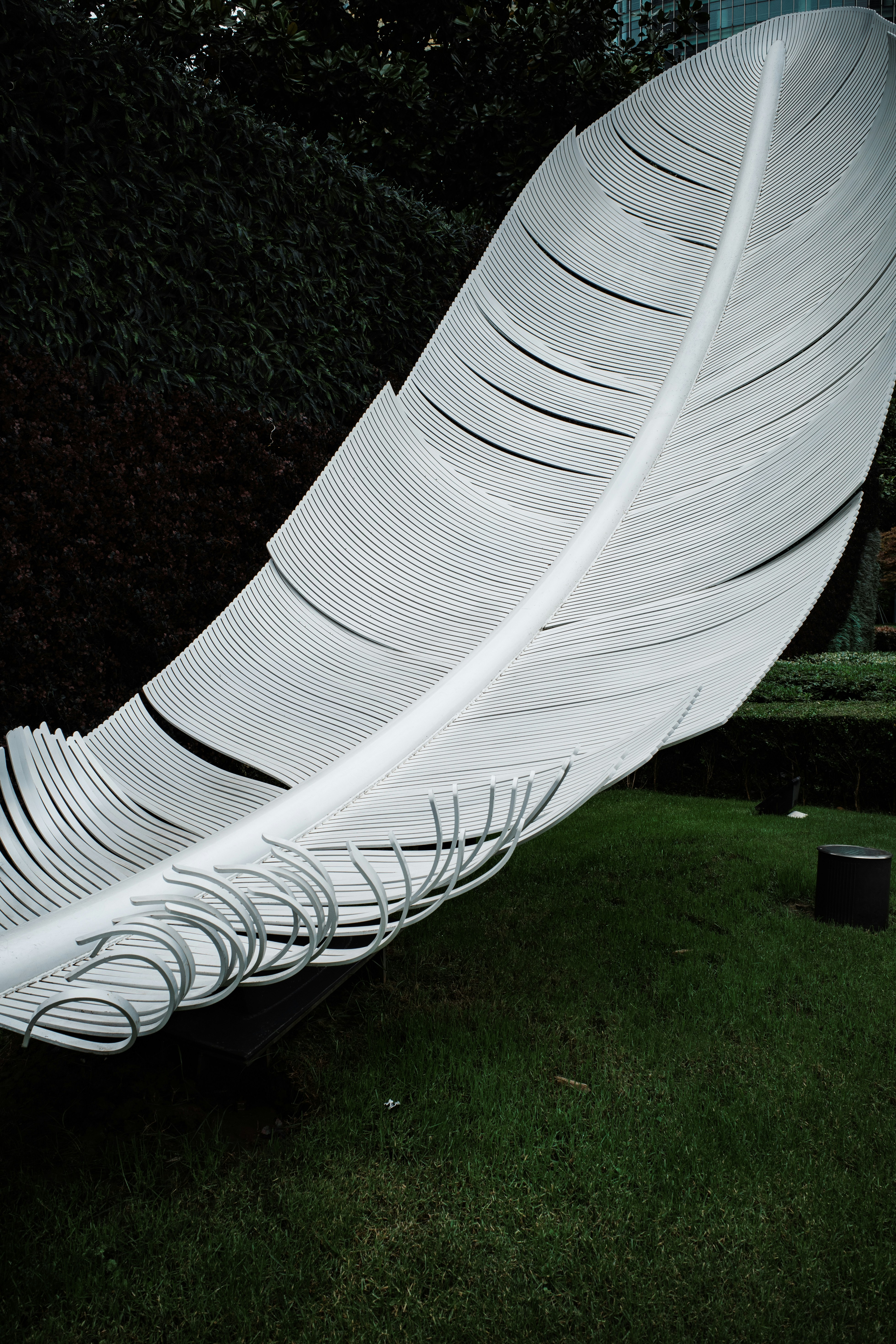 Large white steel sculpture composed of curved slats forms an arch over a grassy lawn, with dark hedges in the background.
