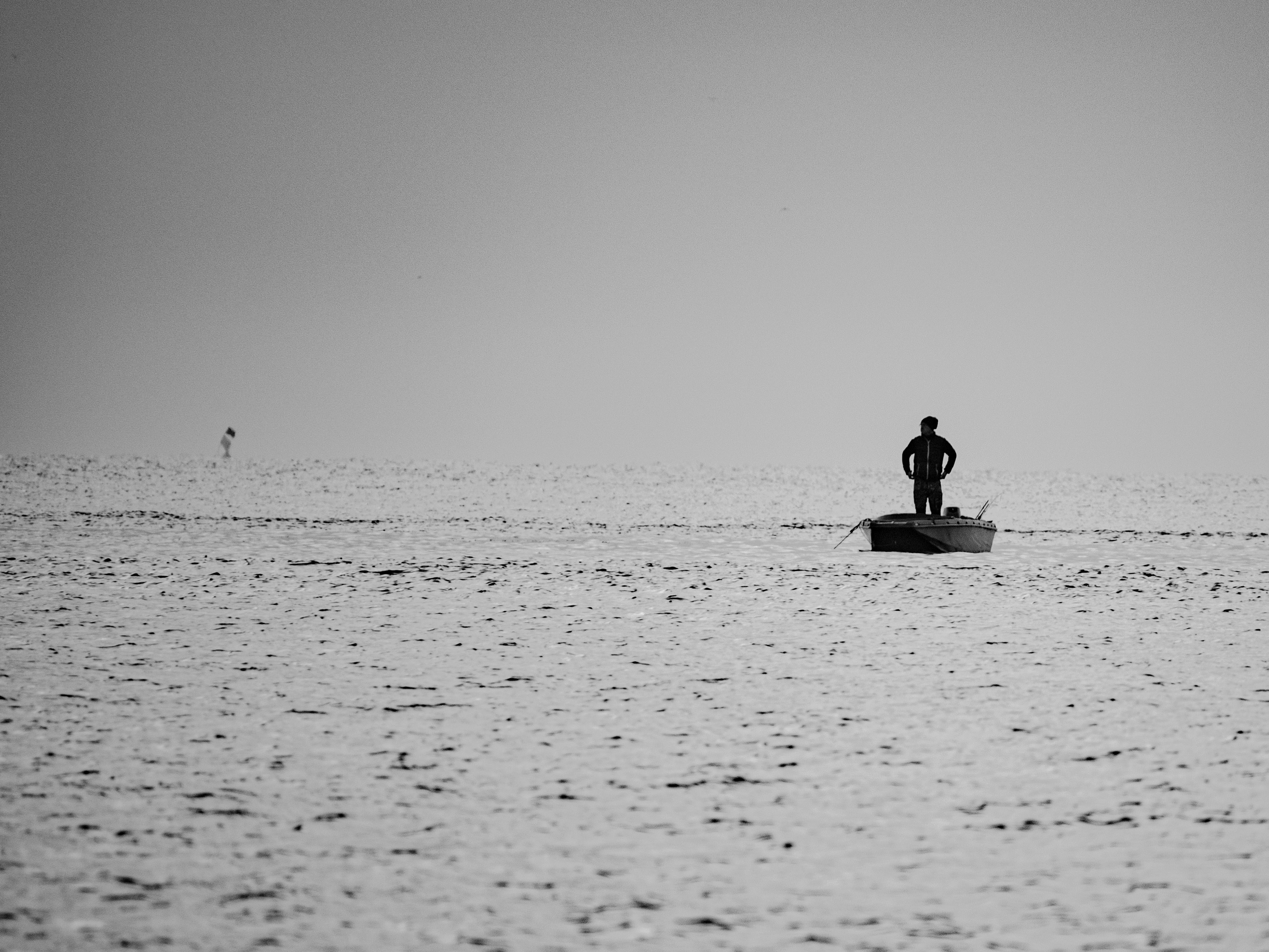 A lone figure stands in a small boat, surrounded by a vast expanse of water under a muted sky. The monochromatic tones enhance the sense of isolation.