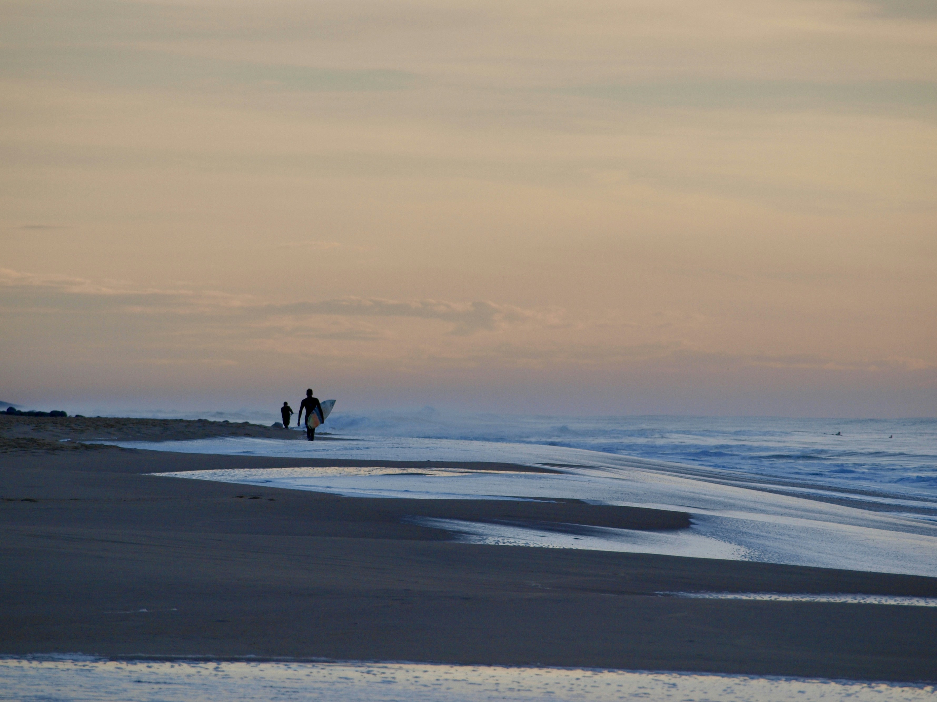 Two surfers walking along a tranquil beach at dusk, with gentle waves lapping at the shore. The soft pastel colors of the sky reflect the serene atmosphere.