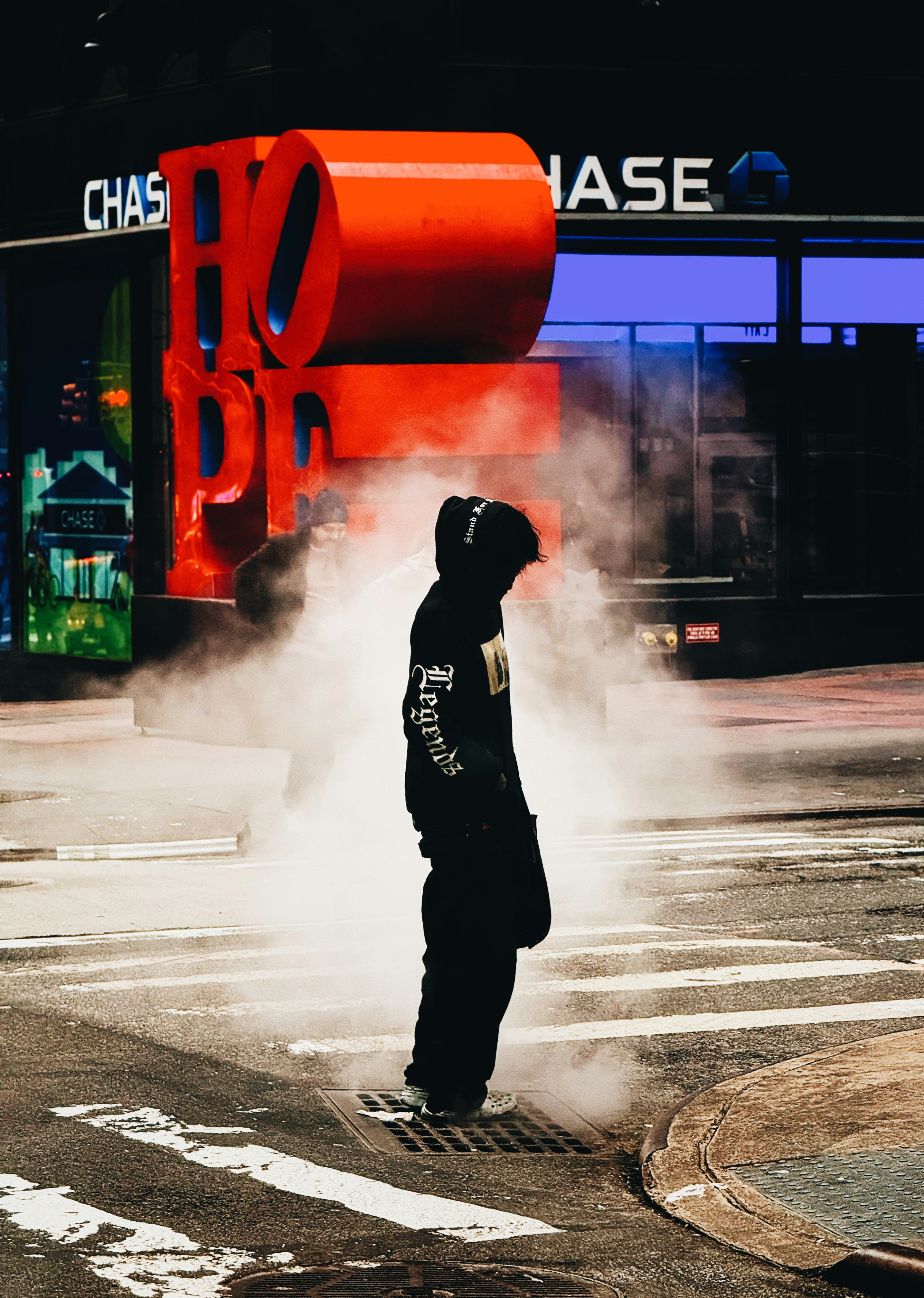 Person in a hoodie stands on a city street surrounded by steam with a large red 'HOPE' sculpture in the background.