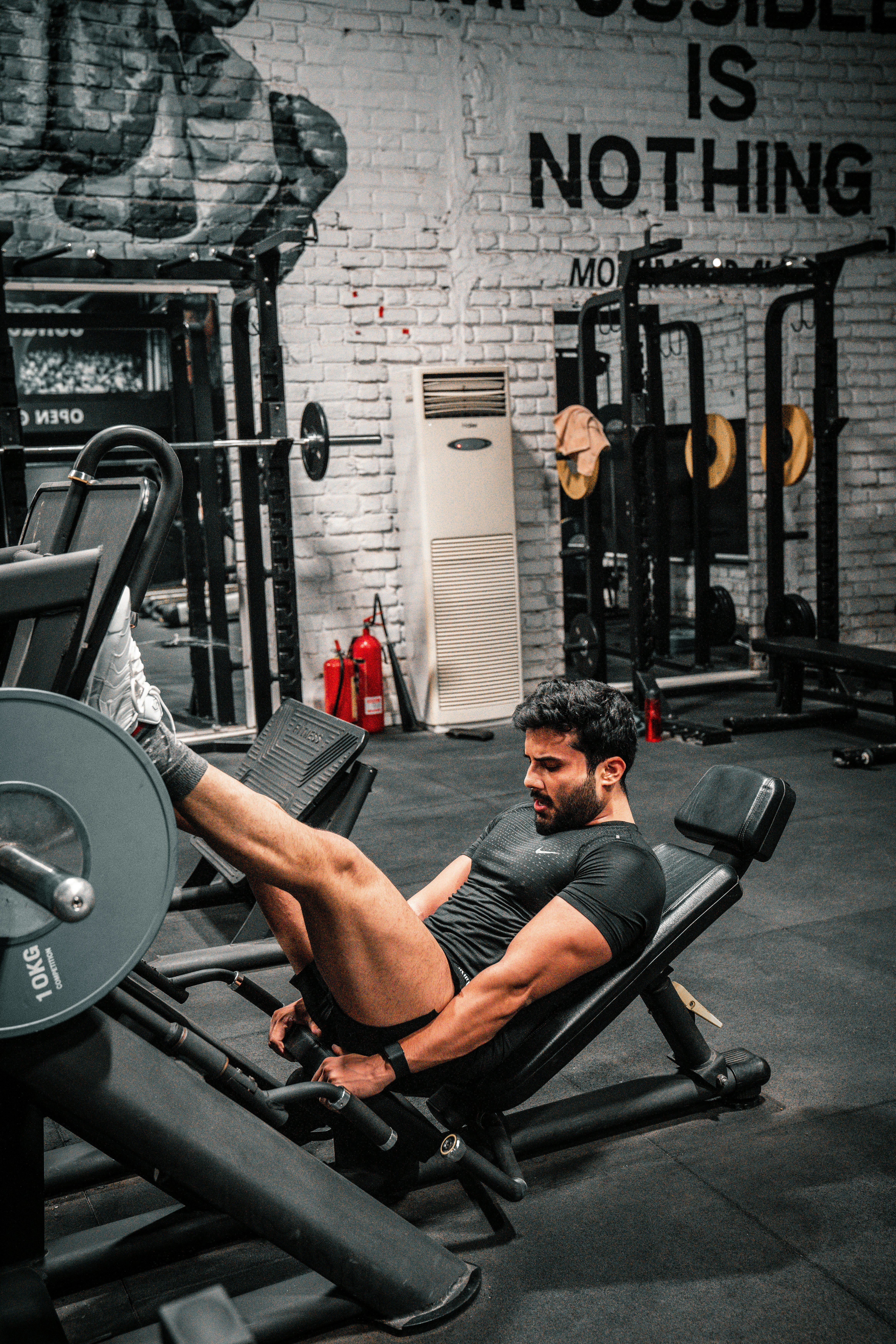 Man engaged in a leg press exercise in a gym, showcasing focus and strength. The backdrop features motivational text on the wall.